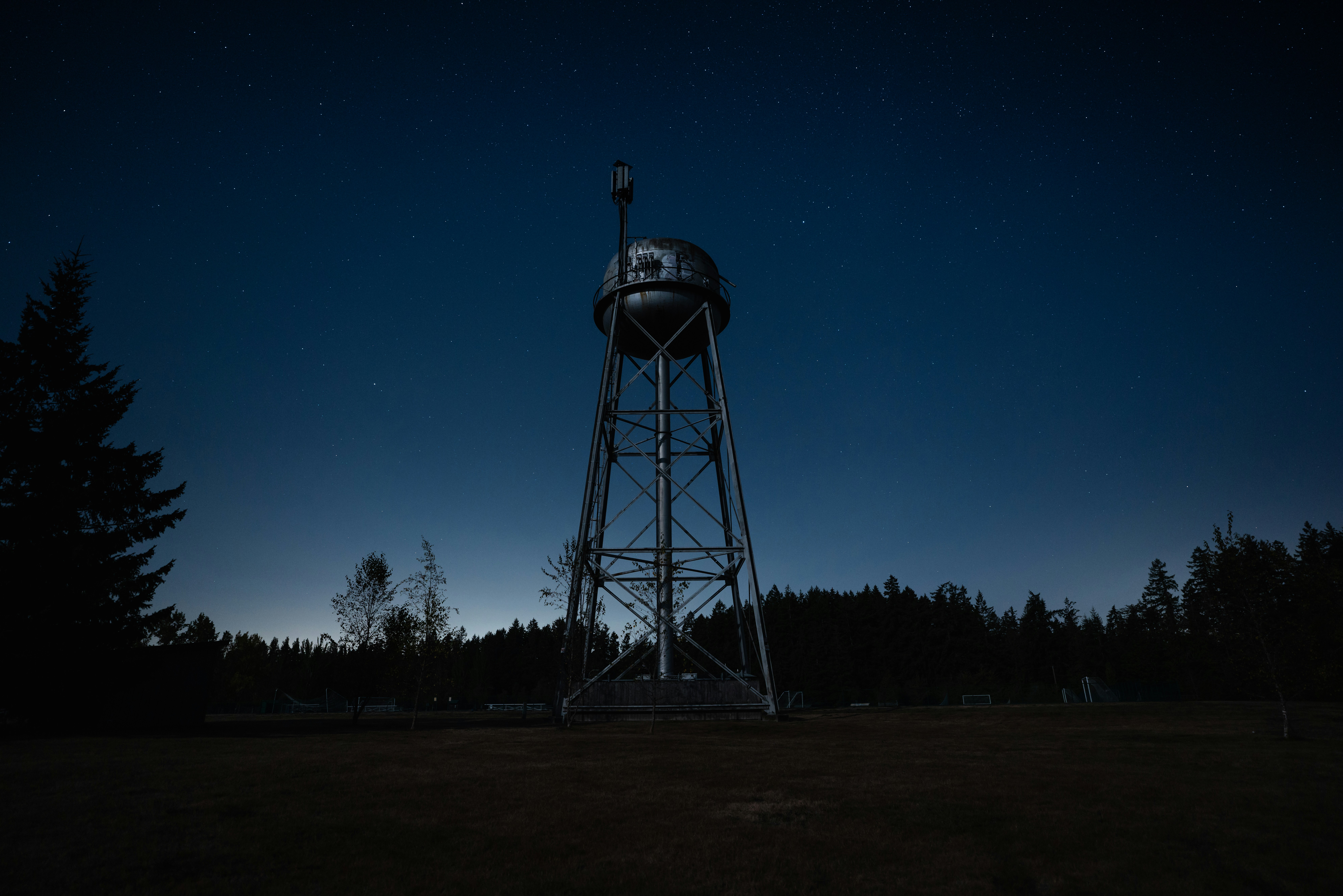 Metal water tower silhouetted against a starry night sky with distant trees lining the horizon.