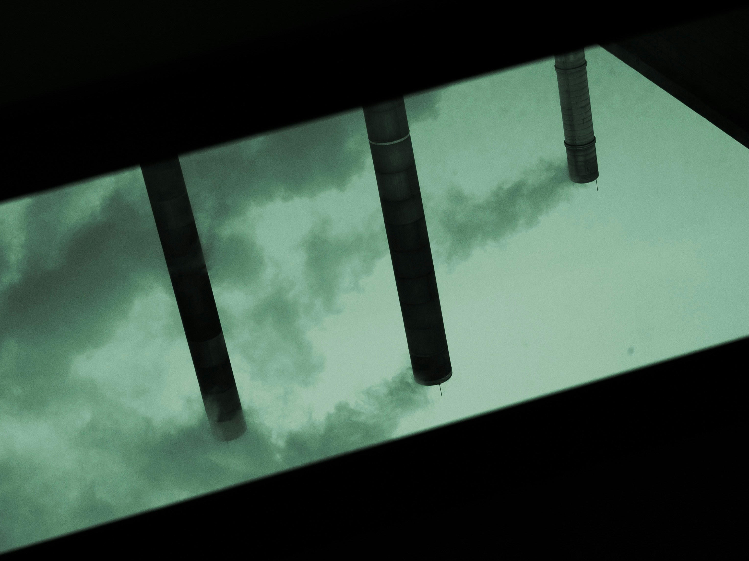 Three smokestacks rise against a muted green sky, casting a stark silhouette that evokes the essence of industrialization. Wisps of smoke intertwine with the clouds above.