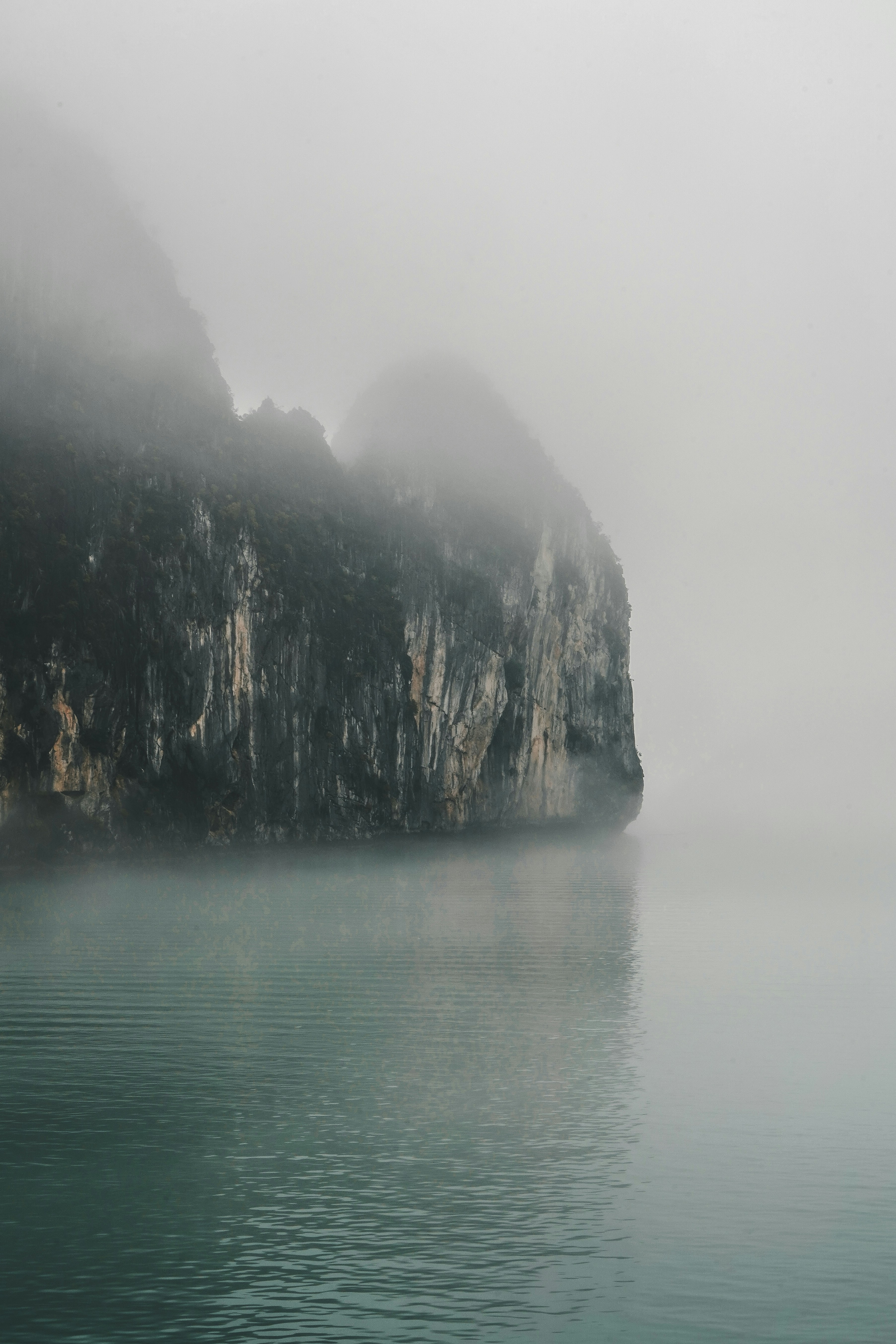 Misty cliffs rise dramatically from tranquil waters, enveloped in a soft fog that blurs the horizon. The scene evokes a sense of calm and mystery.