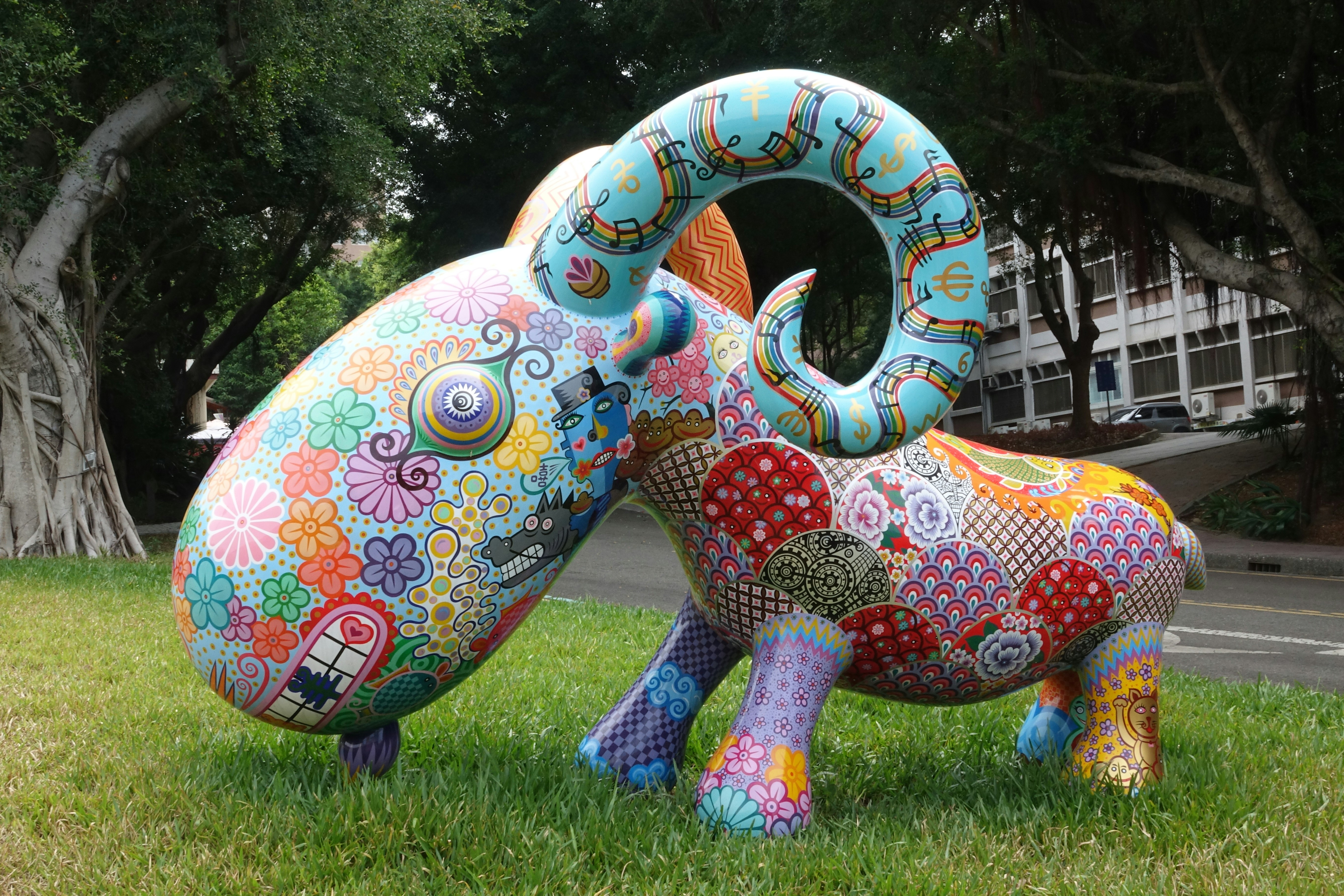 Colorful sculpture of a patchwork-patterned animal with large spiral horns on a grassy area.