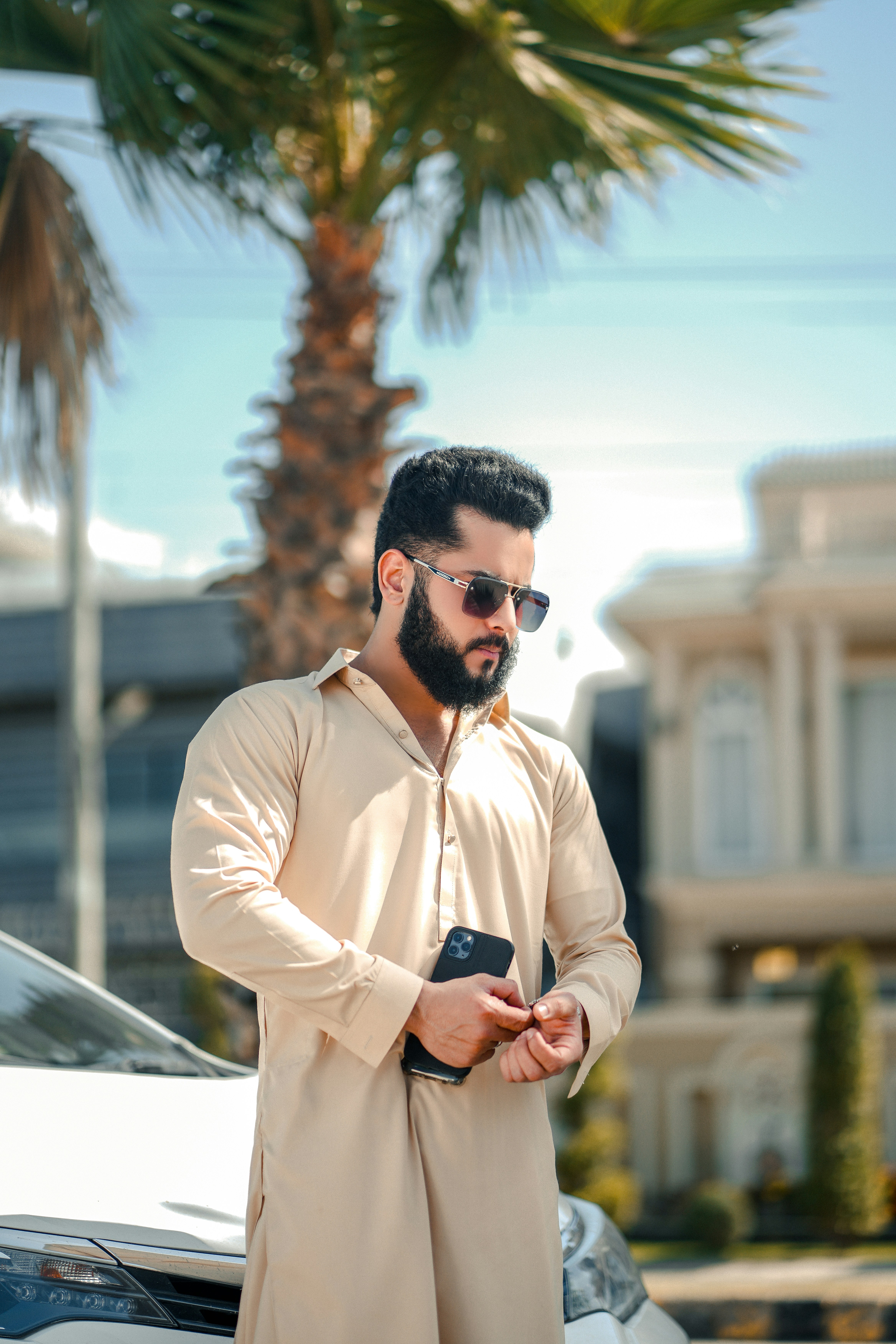 Man in traditional attire standing by a car under palm trees on a sunny day.