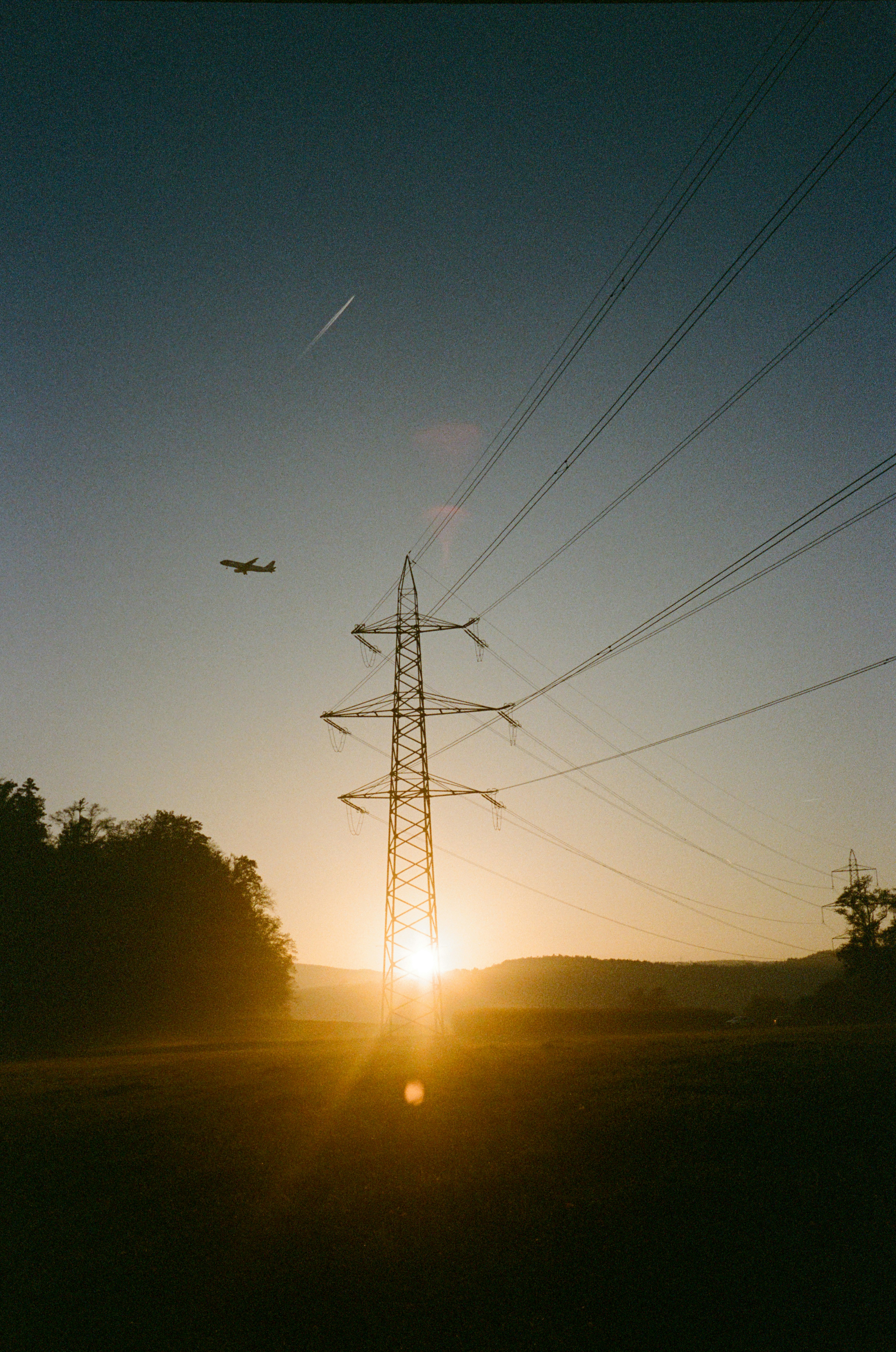 Airplane flying over a silhouetted power line tower at sunset with a clear sky.