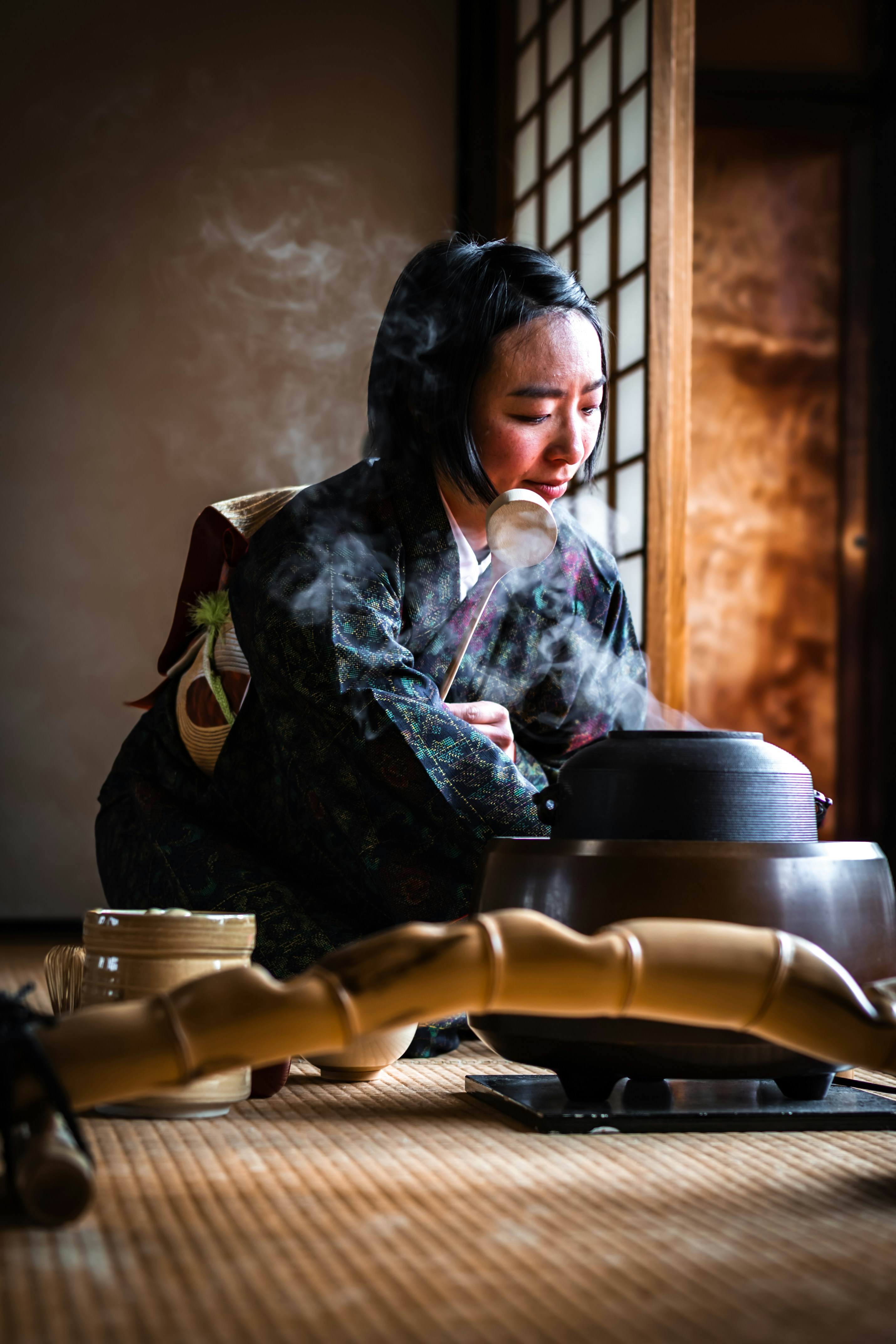 Woman in traditional kimono tending to a steaming pot in a serene tea room, surrounded by bamboo and tea utensils.