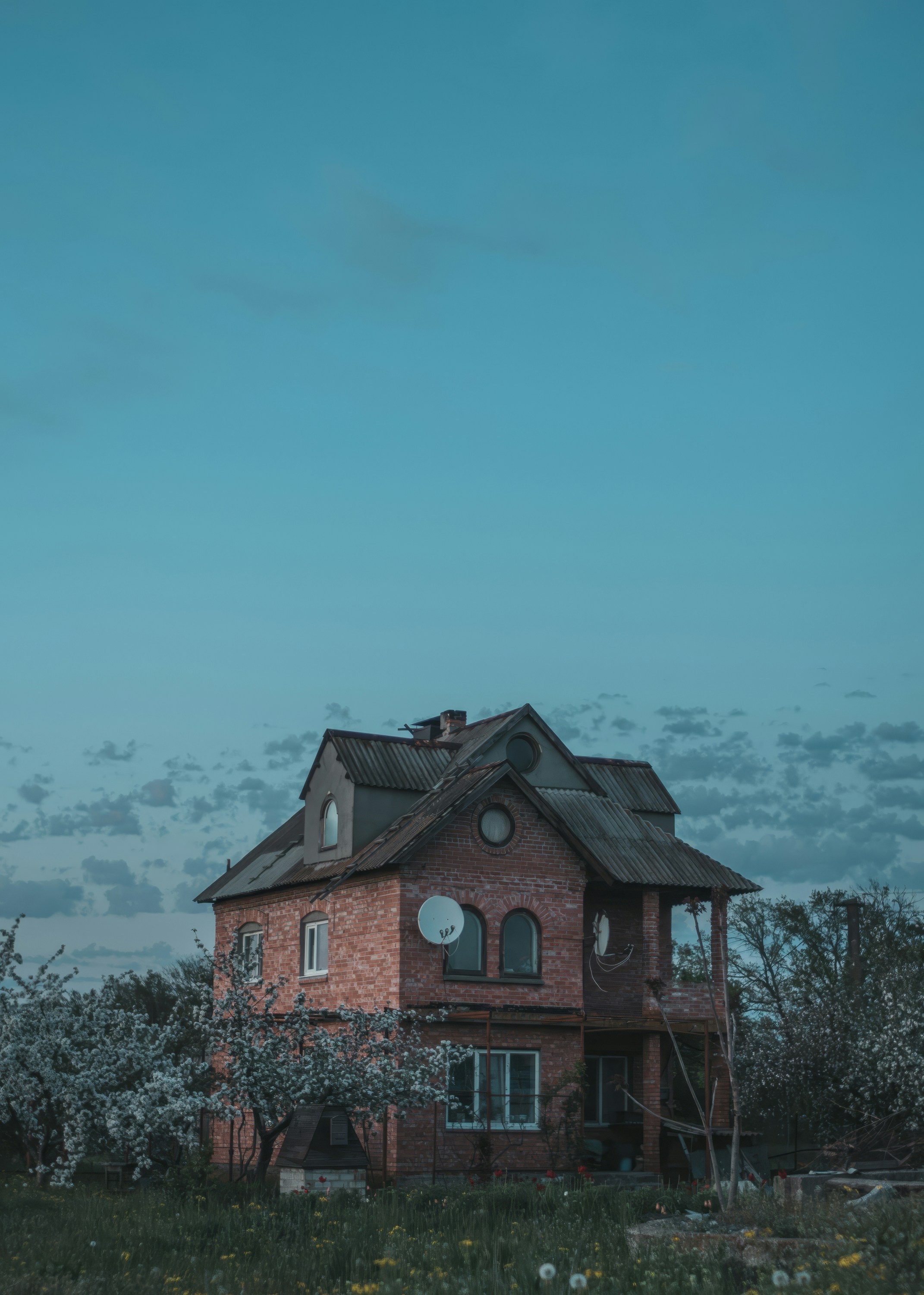A brick house with a satellite dish and multiple windows stands in a field surrounded by blooming trees under a blue sky with scattered clouds at dusk. The unique architectural design features both rectangular and circular windows.