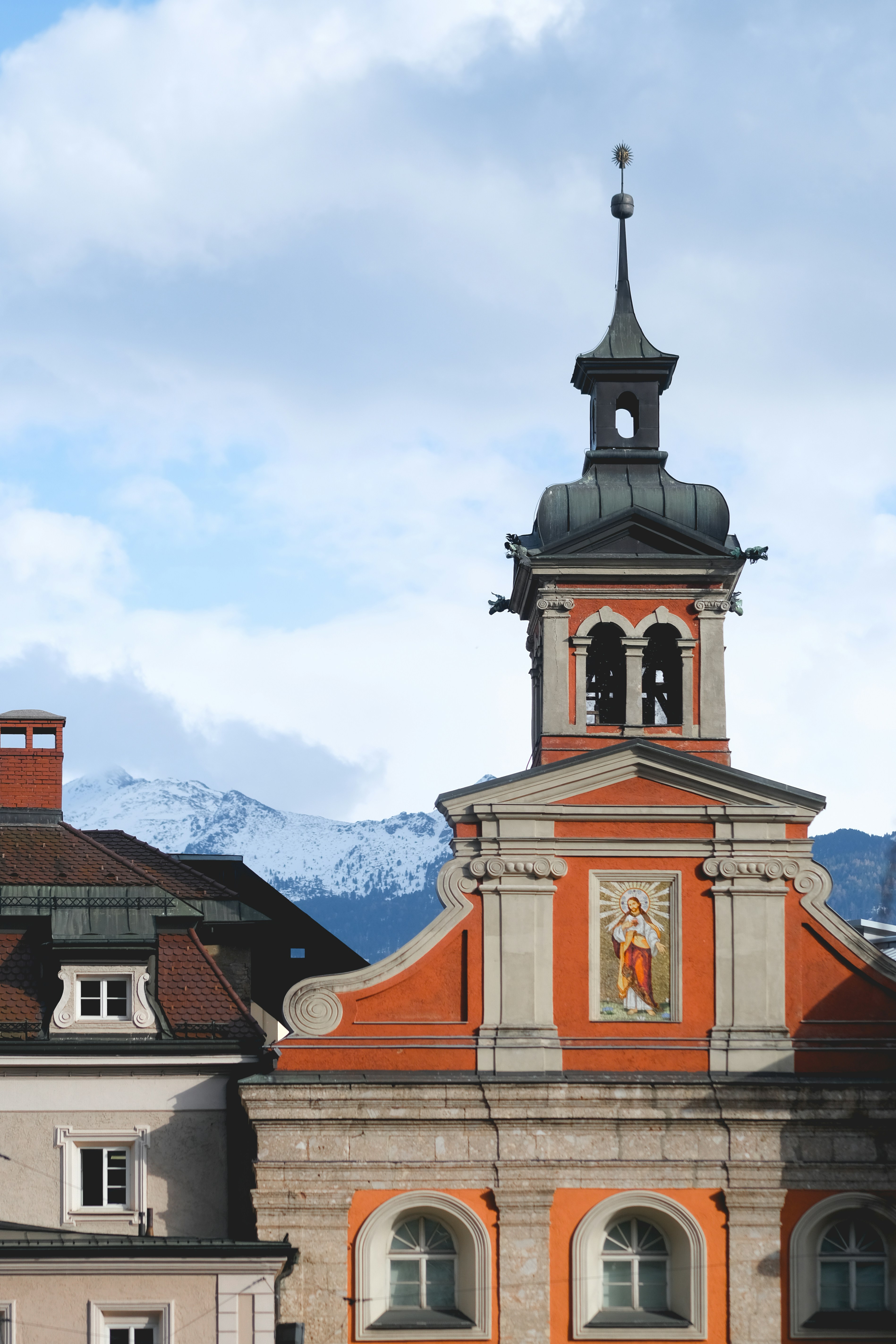 European church tower with ornate spire and religious fresco set against snow-capped mountains and vibrant orange facade.