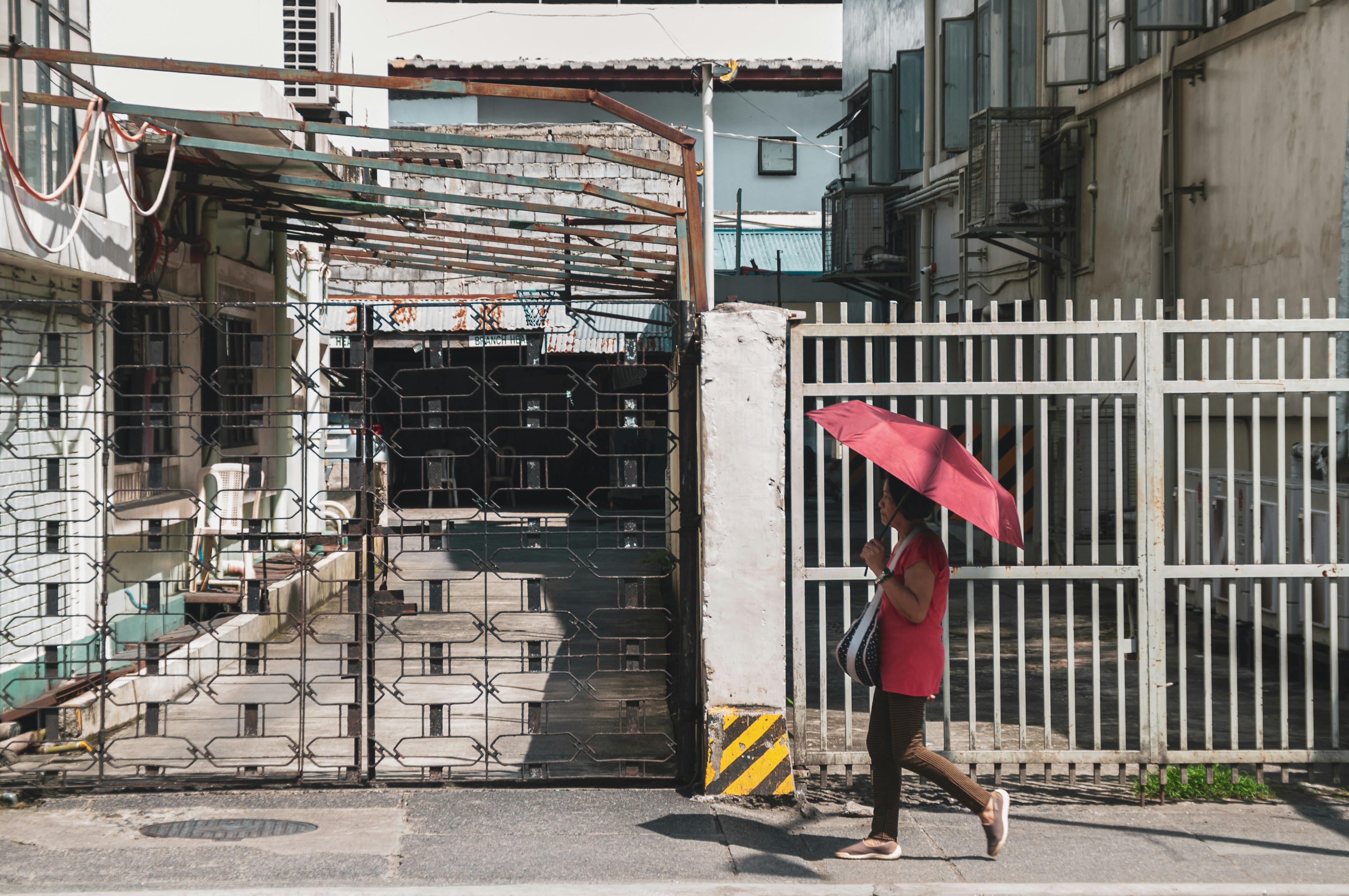 Person walking past industrial buildings holding a red umbrella under the sun.
