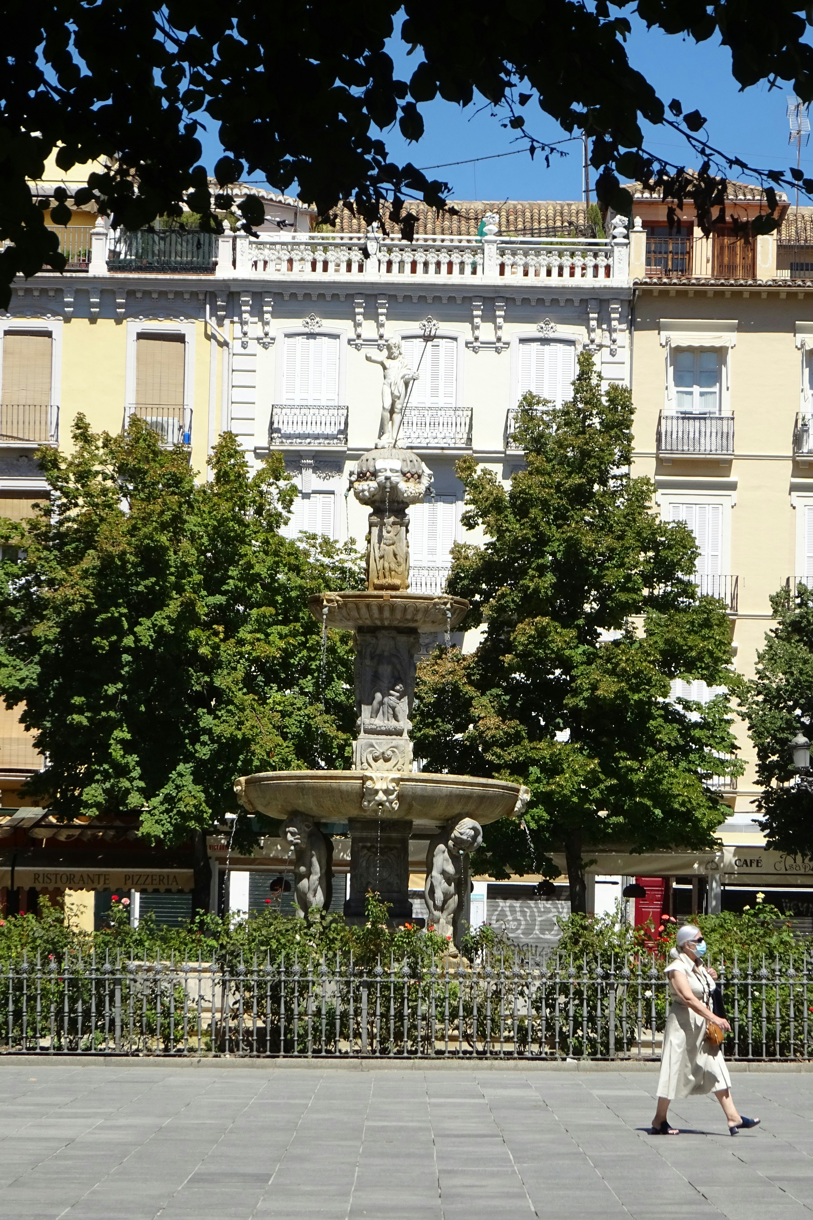 A classical multi-tier fountain stands at the center of a sunlit plaza, framed by trees and pastel European buildings. A lone pedestrian walks by, adding a sense of scale to the urban scene.