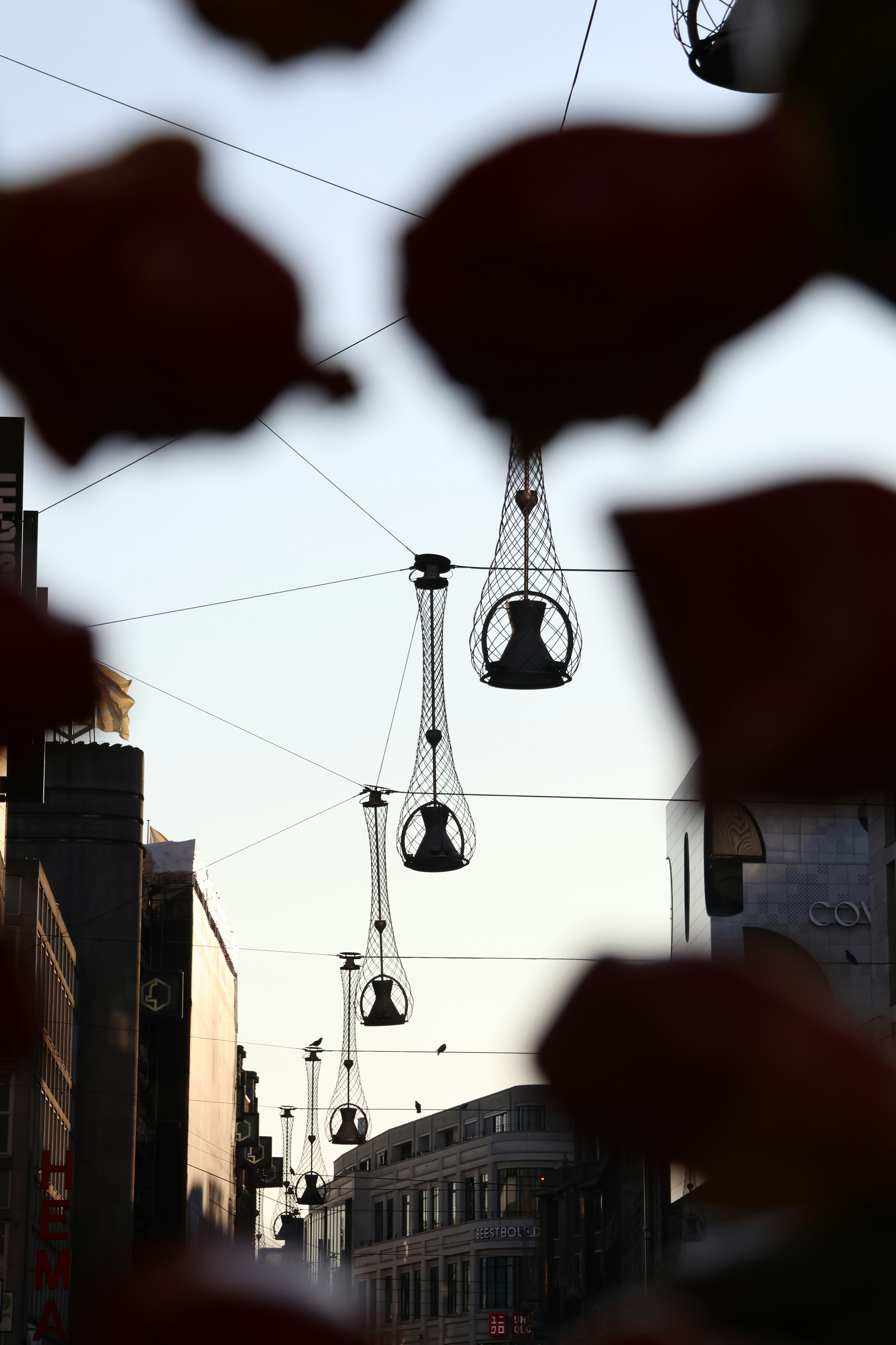 Unique street lamps hanging above a bustling urban street, framed by blurred foliage in the foreground.