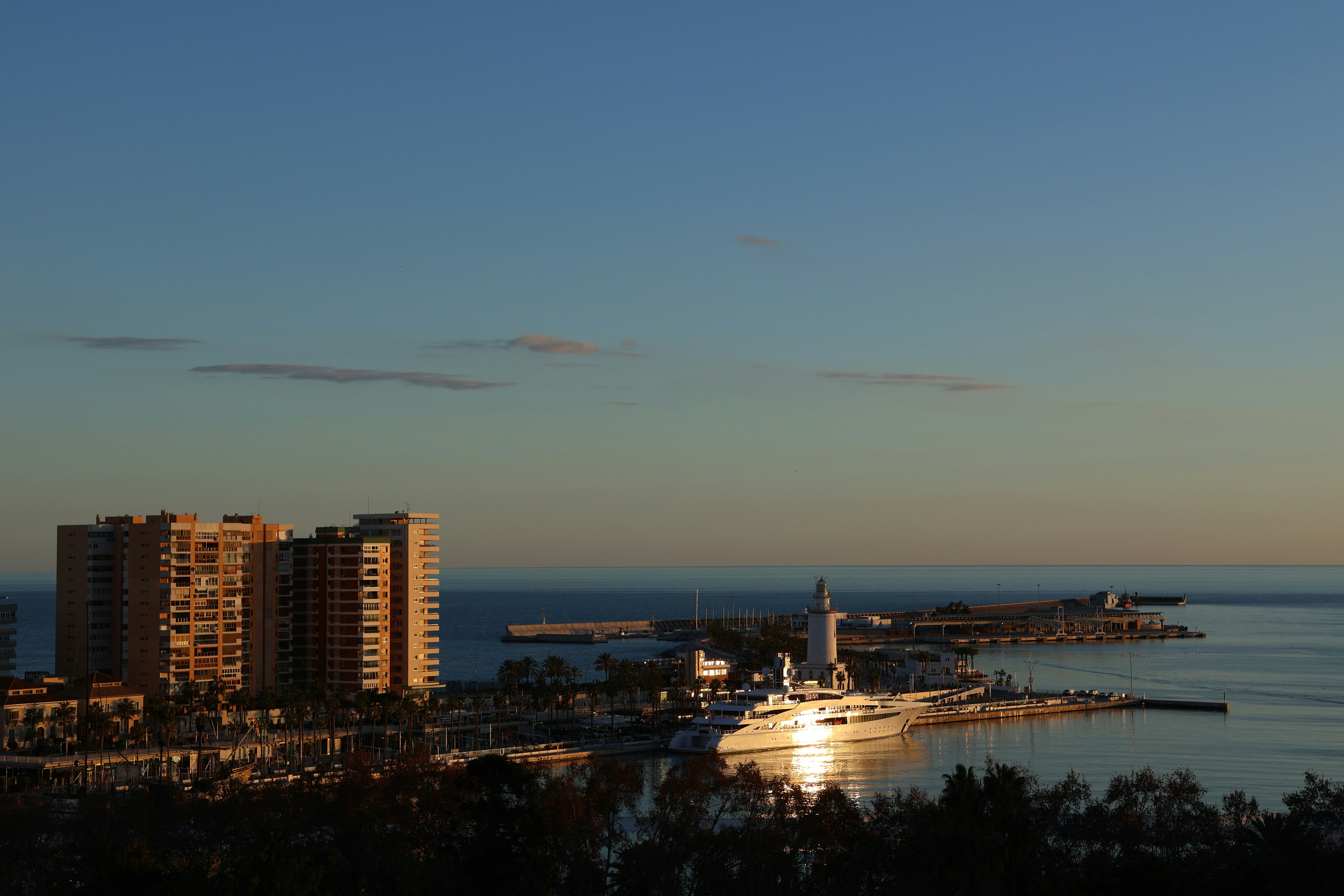 Sunset casts a warm glow over a coastal cityscape and harbor with docked boats.