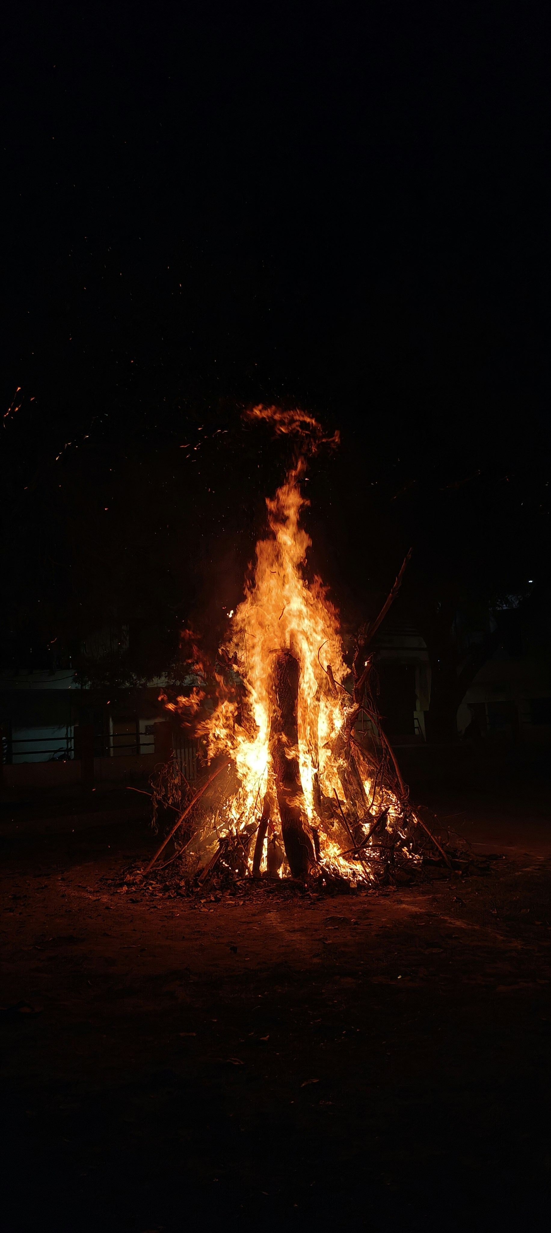 A vibrant bonfire crackles under the night sky, illuminating the surrounding darkness with warm hues. Sparks dance upwards, creating a mesmerizing display.