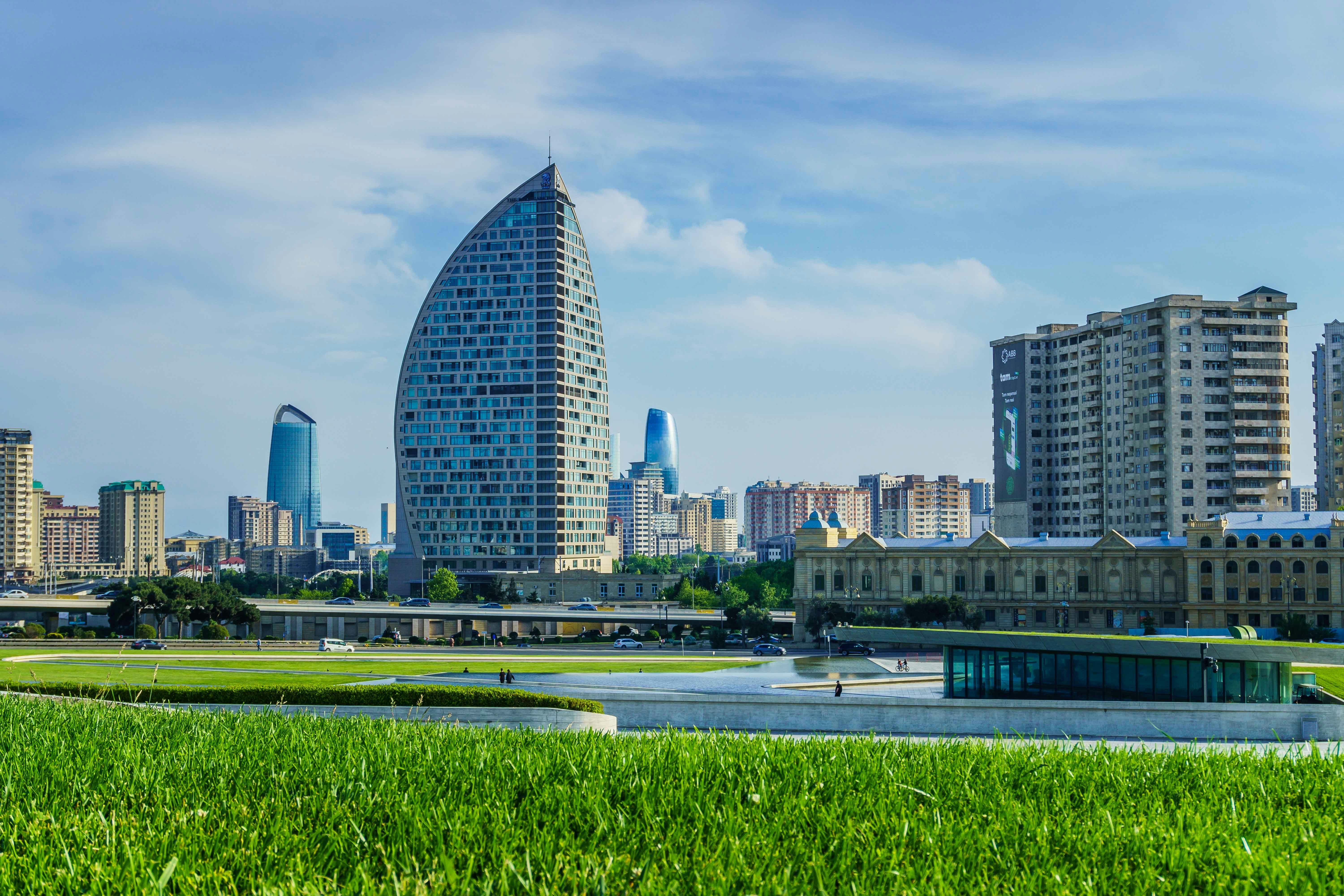 Modern skyscrapers rise behind a lush green foreground under a clear blue sky.