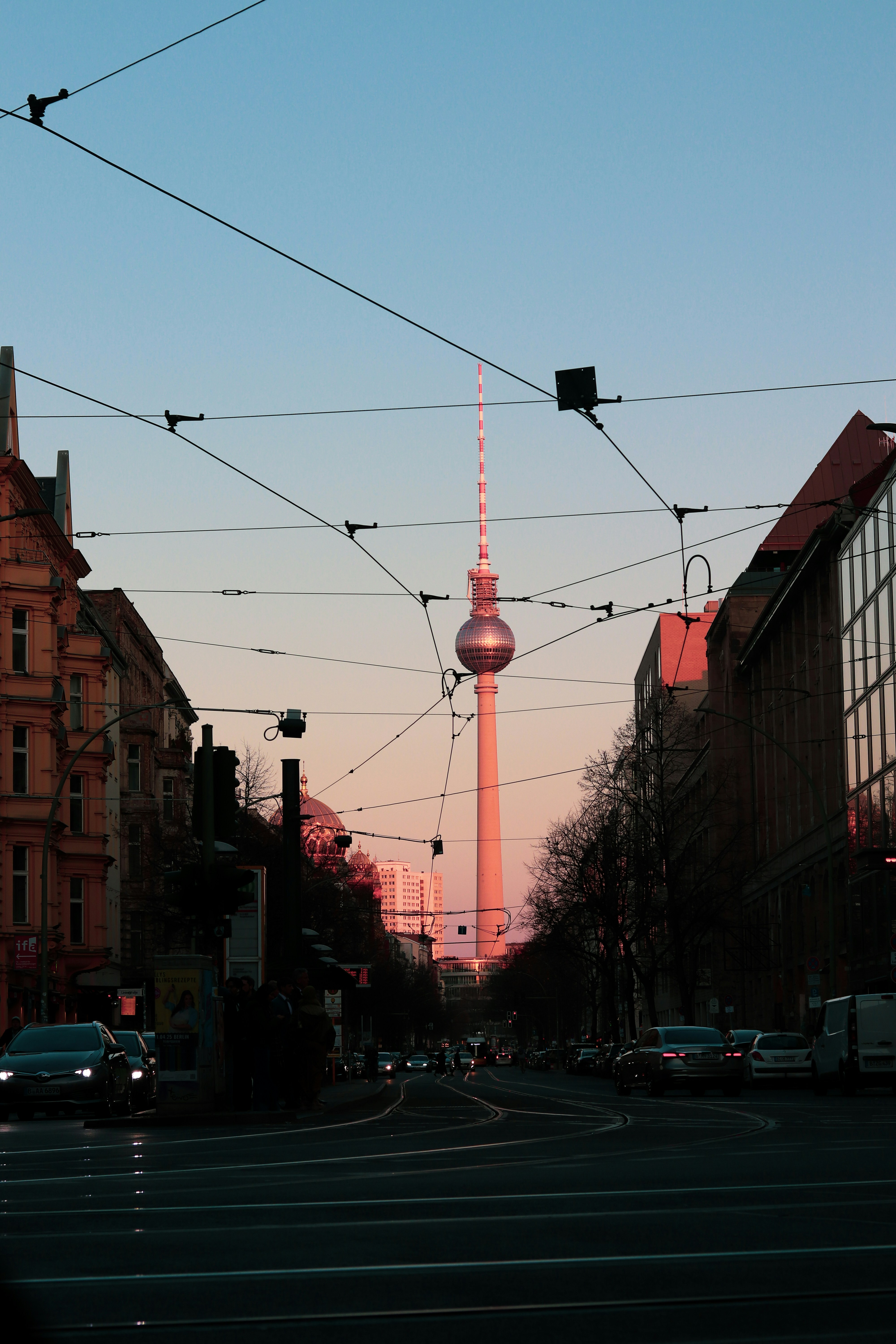 The Berlin TV Tower rises prominently against a pastel sky, framed by urban architecture and tram lines. A busy street scene captures the essence of city life.