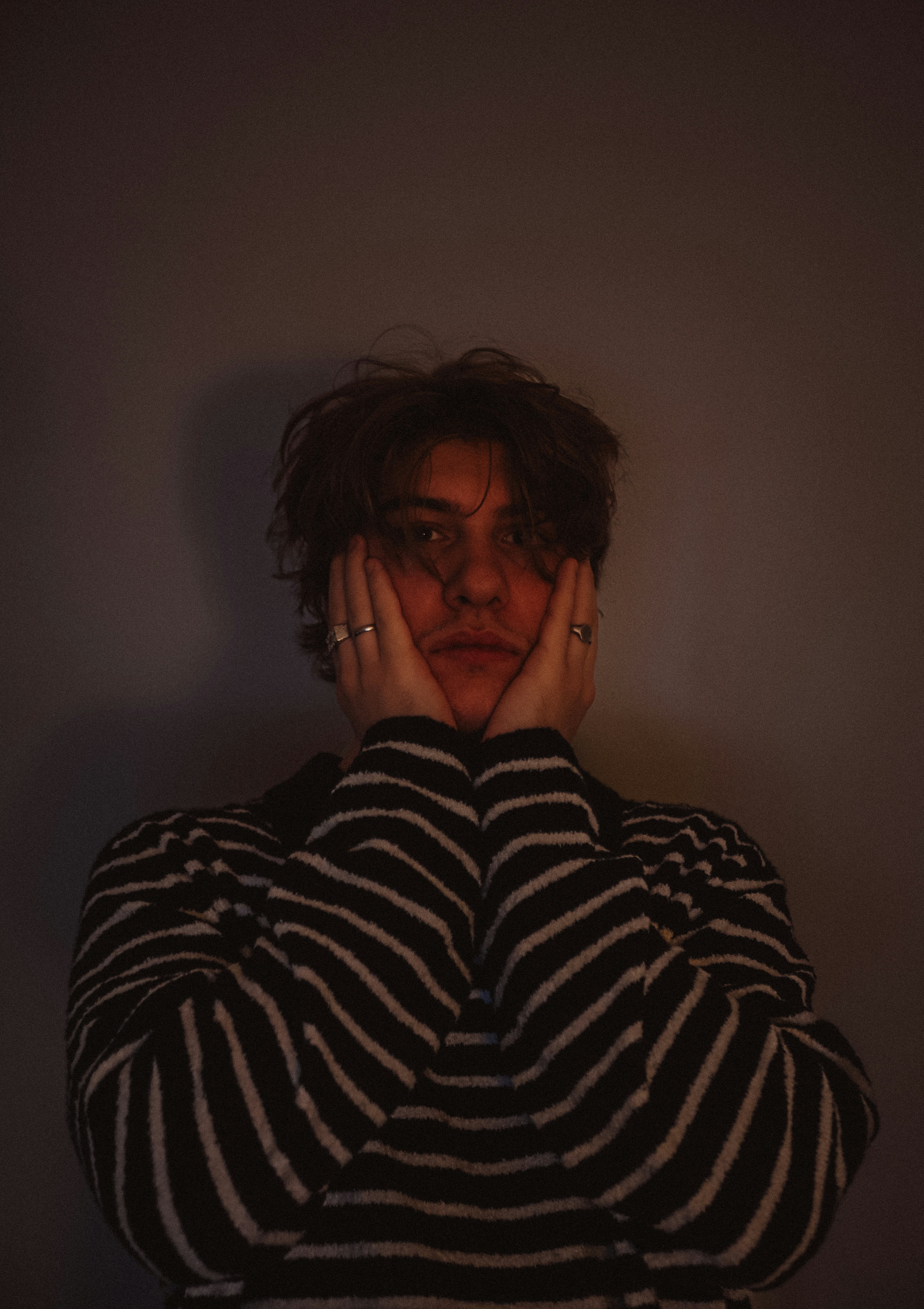 Young person with tousled hair and striped sweater resting hands on face, against a muted background. The image conveys a sense of introspection.