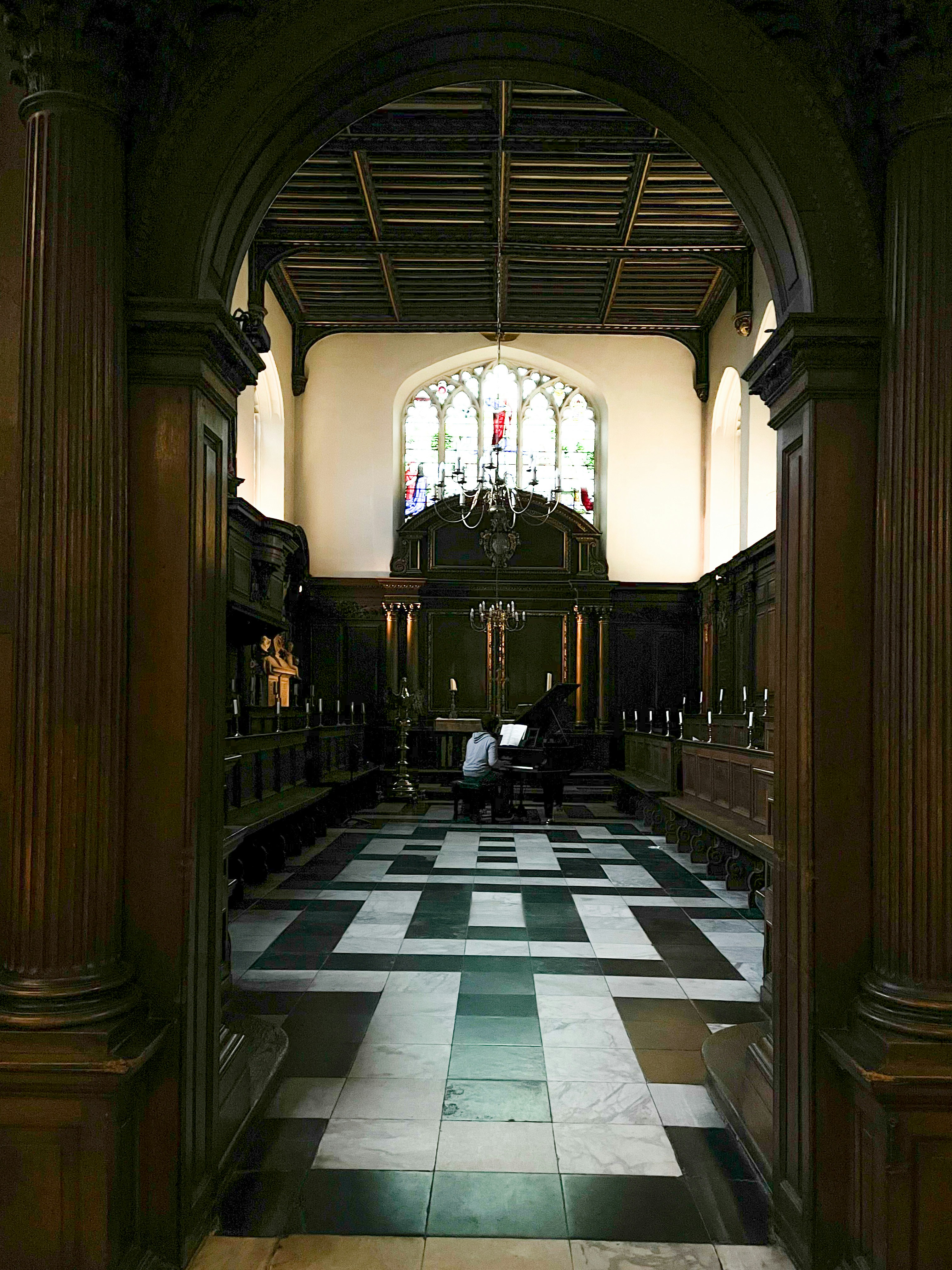 Gothic archway frames a dimly lit chapel interior with checkerboard floor and stained glass window.