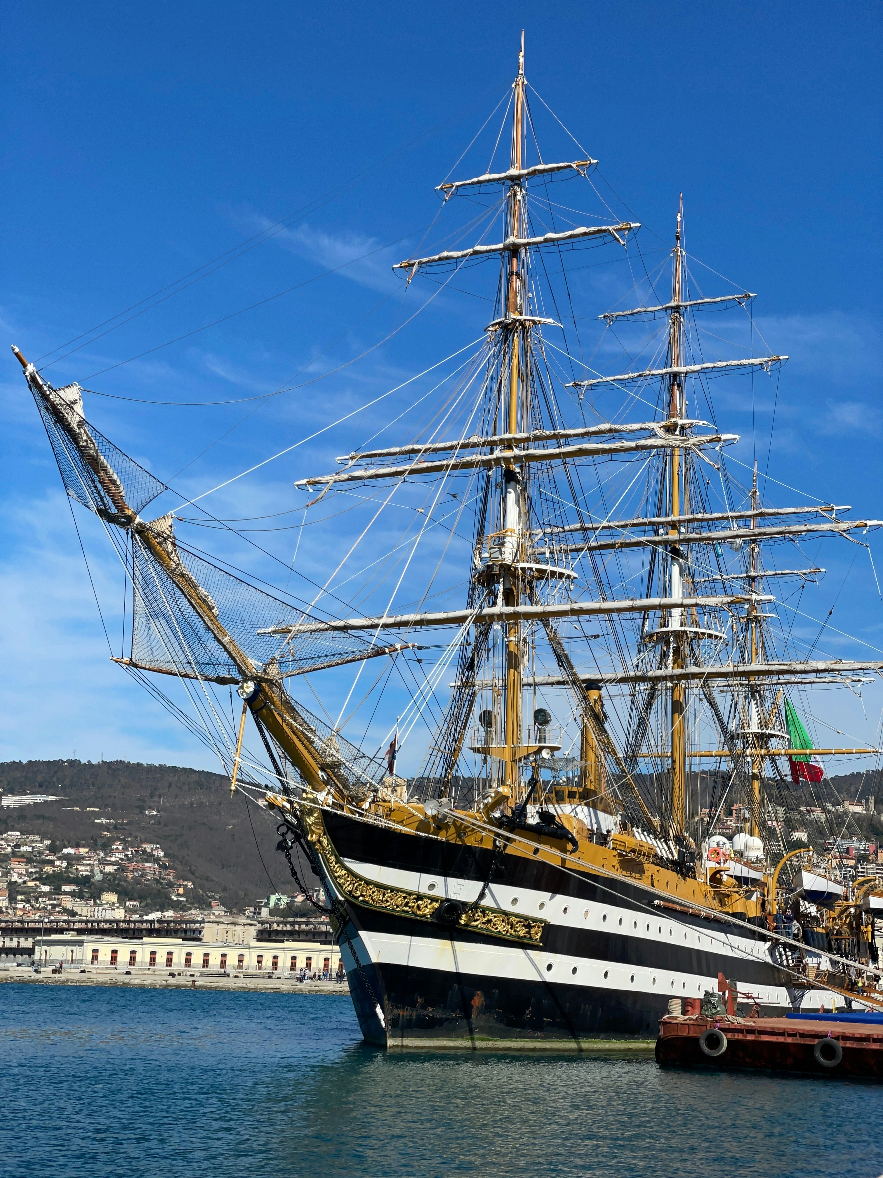 Tall ship anchored in a harbor with three masts and rigging against a backdrop of hills and blue sky.