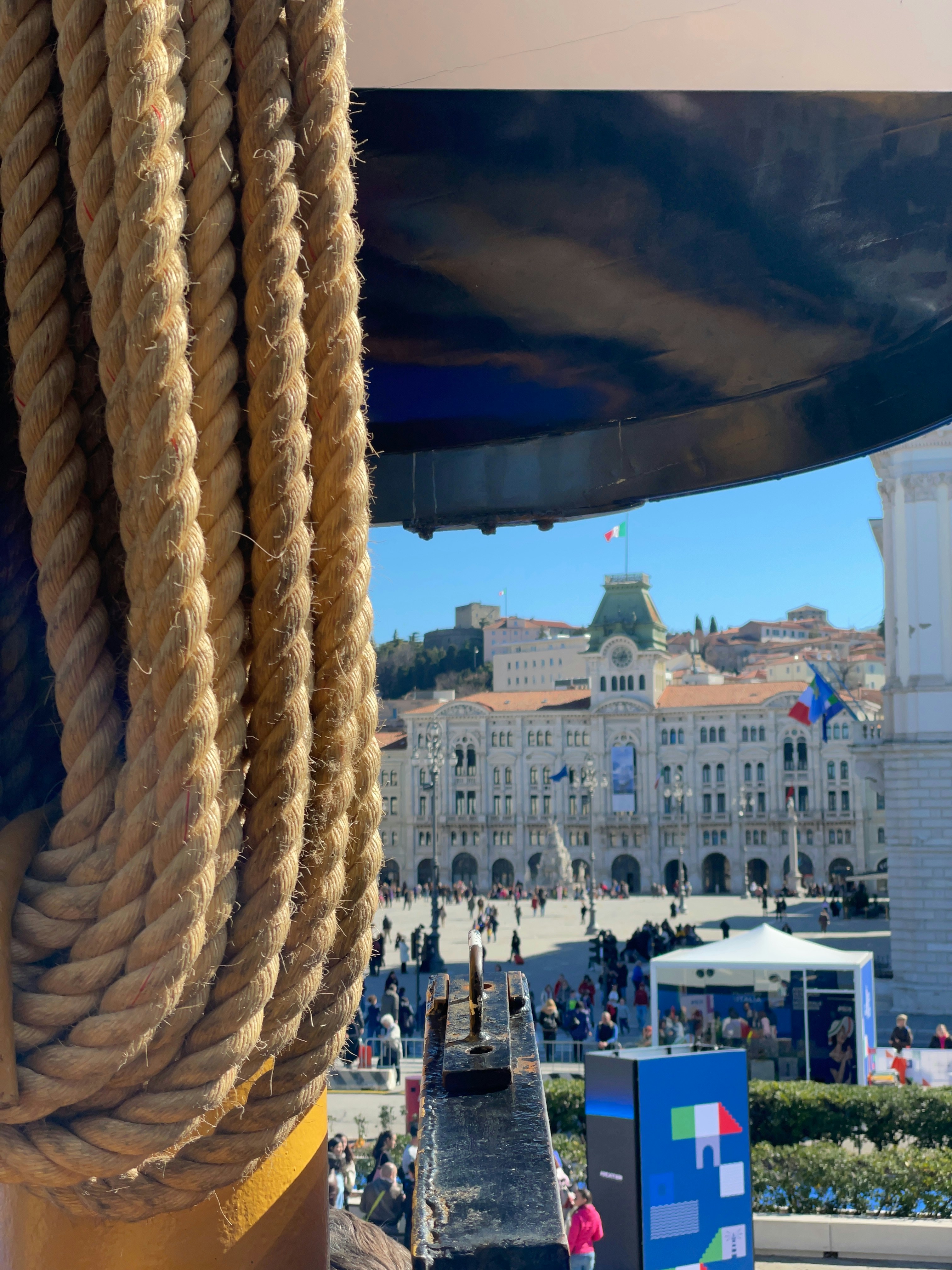 Thick ship ropes frame a bustling plaza with historic architecture under a clear blue sky.