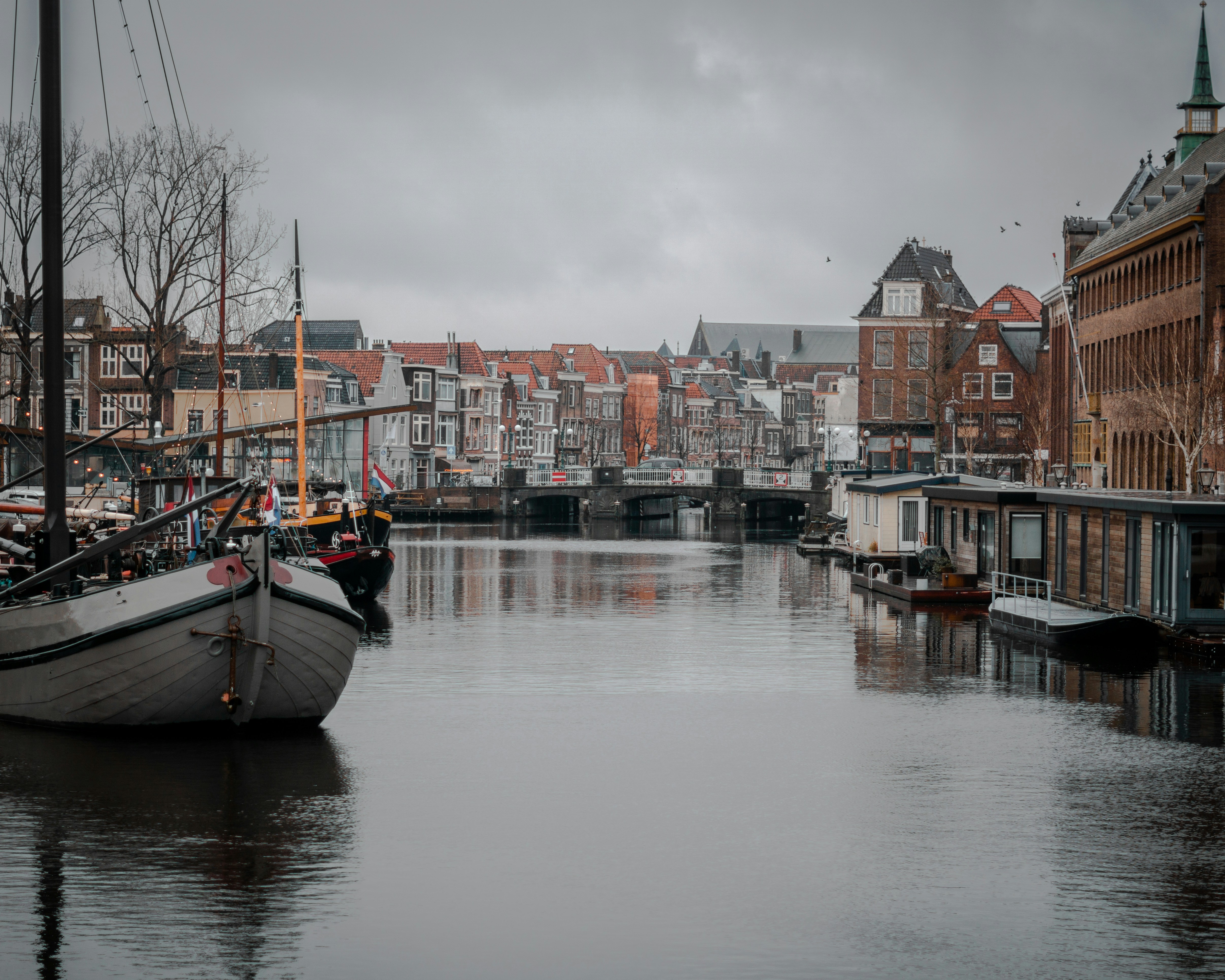 Overcast sky reflecting on a calm canal lined with moored boats and historic buildings.