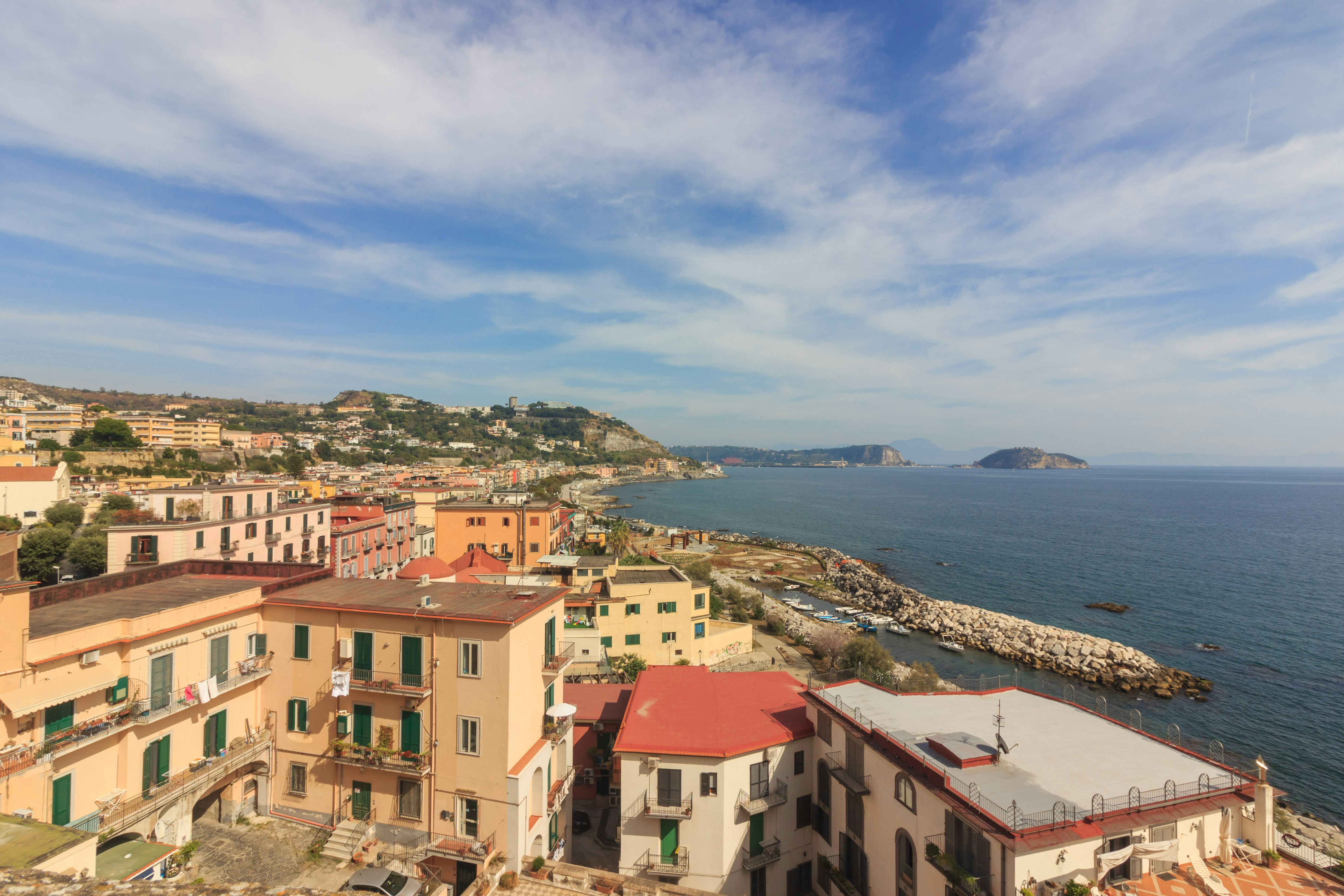 Pastel-colored buildings overlooking a rocky coastline with distant islands under a blue sky.