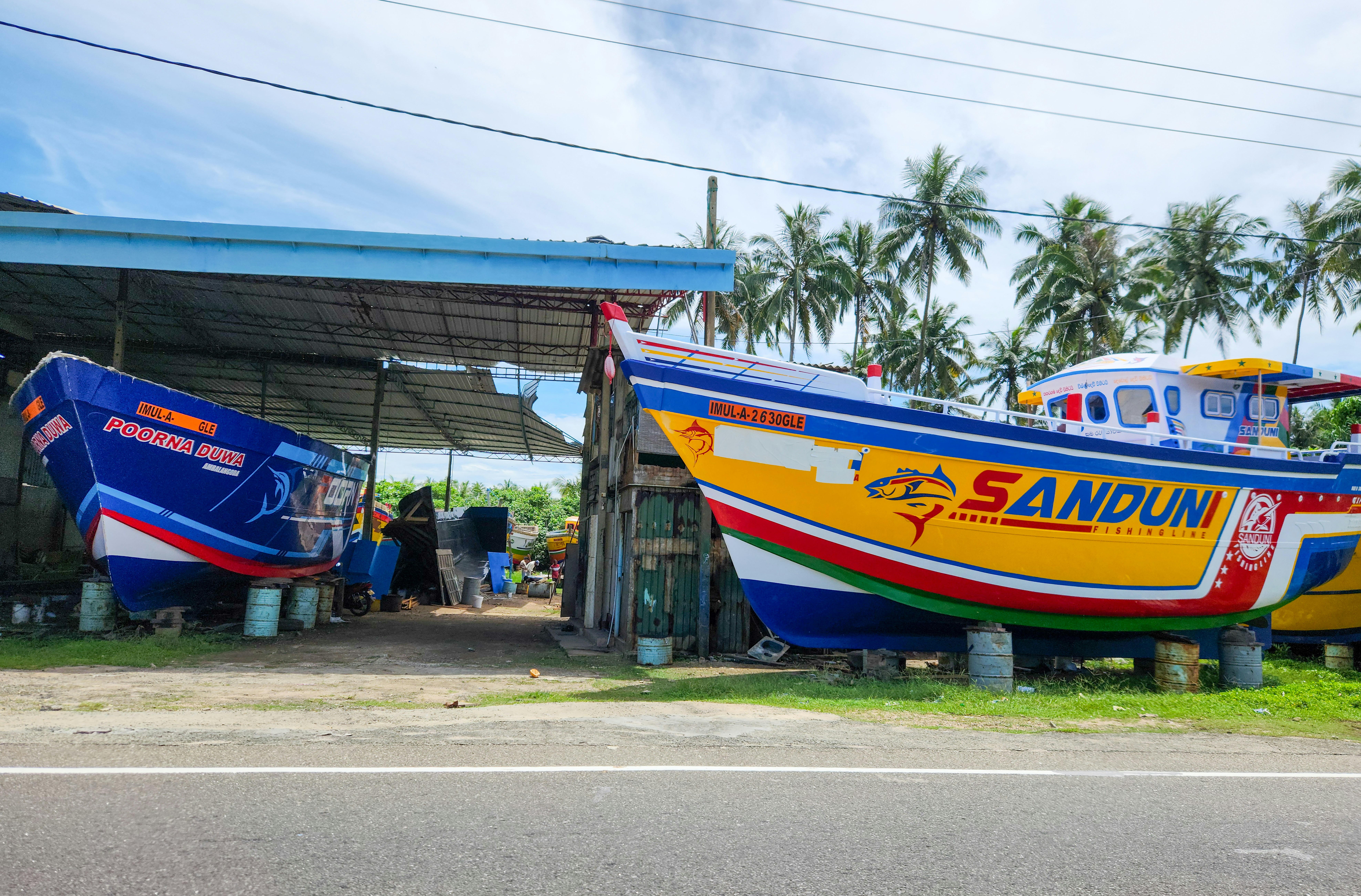 Two colorful Sri Lankan fishing boats resting ashore on stands in a boatyard or coastal area for maintenance.