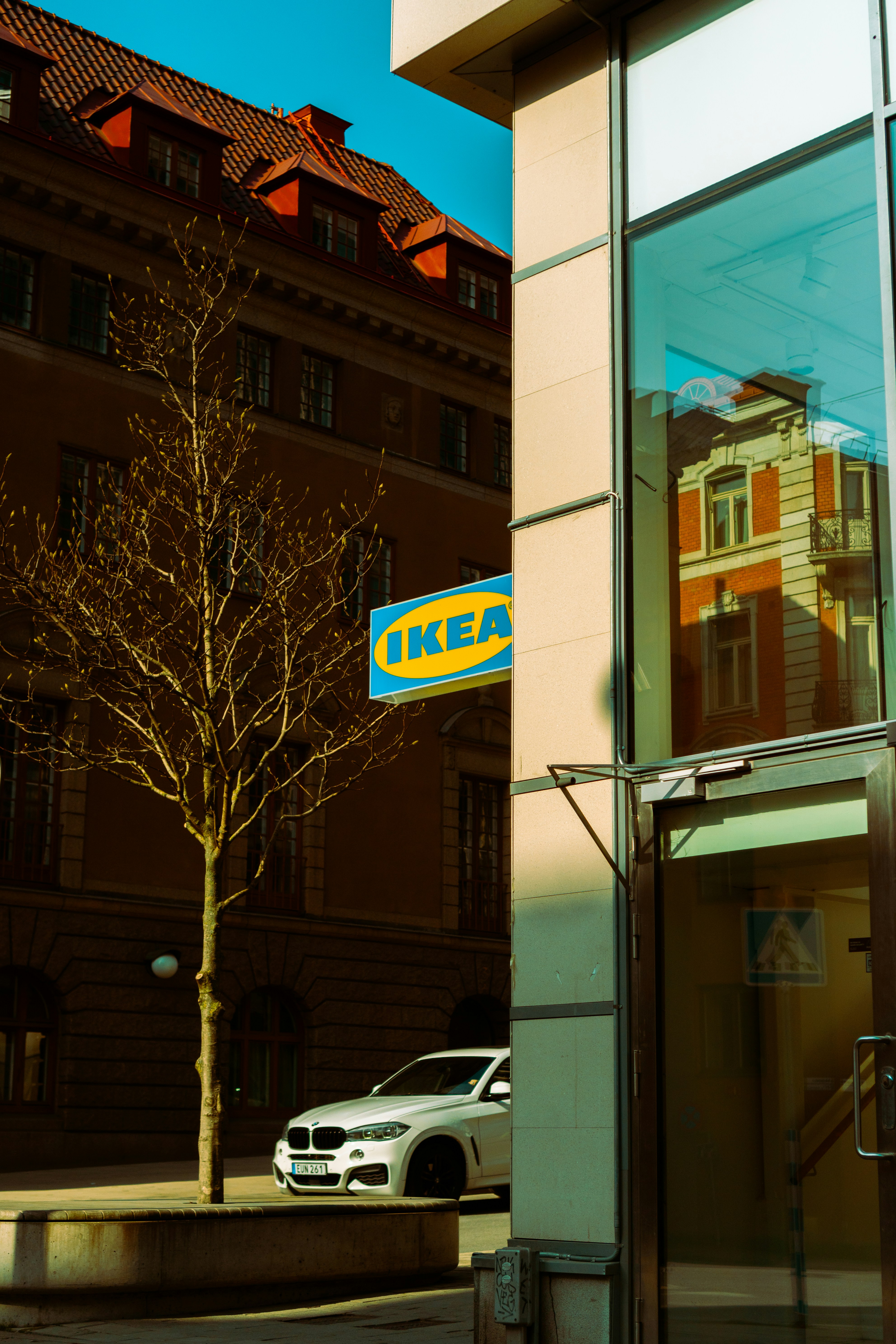 Glass facade reflecting historic architecture with an IKEA sign and parked car in the background.