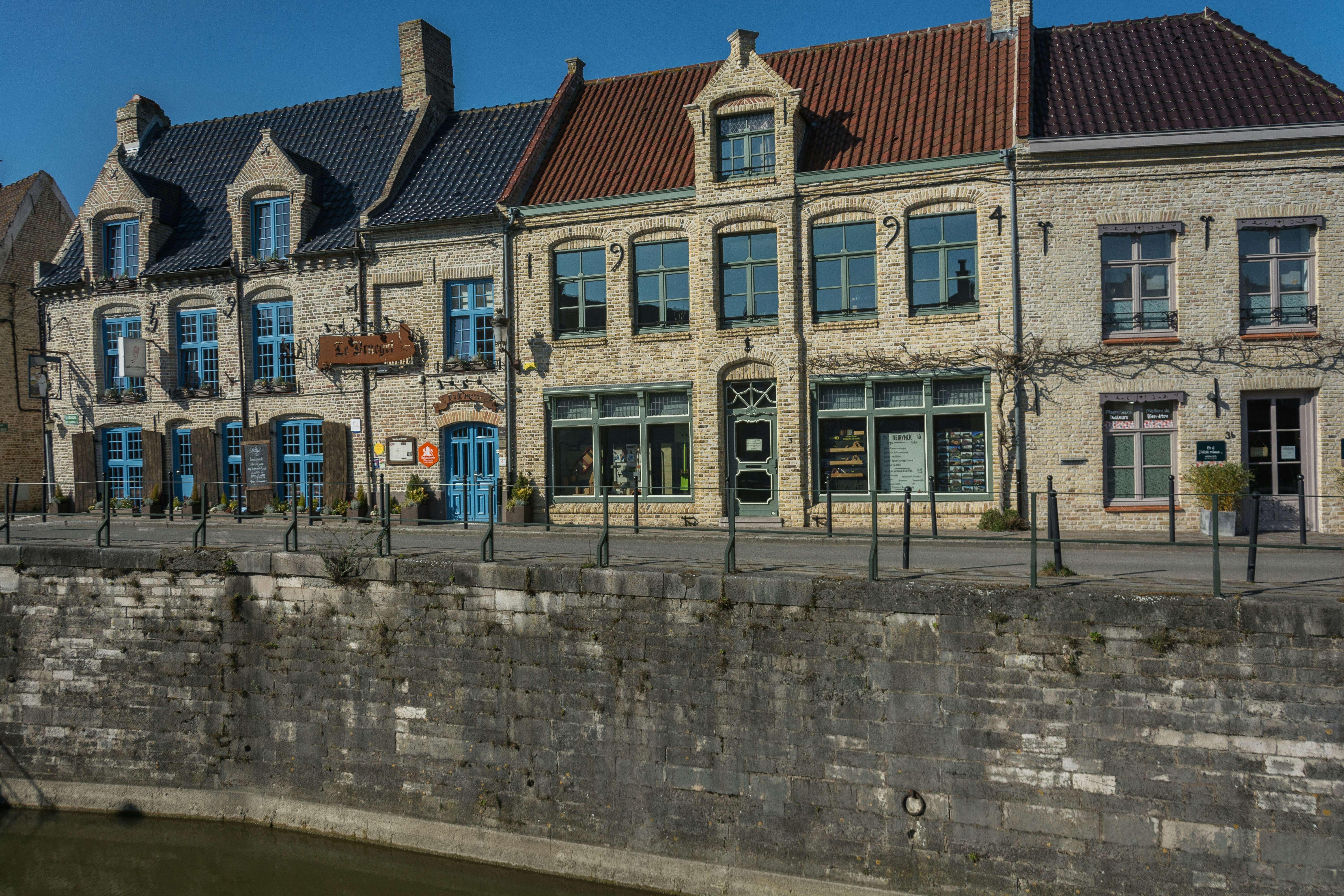 Row of traditional stone buildings with colorful shutters along a riverside embankment under a clear blue sky.