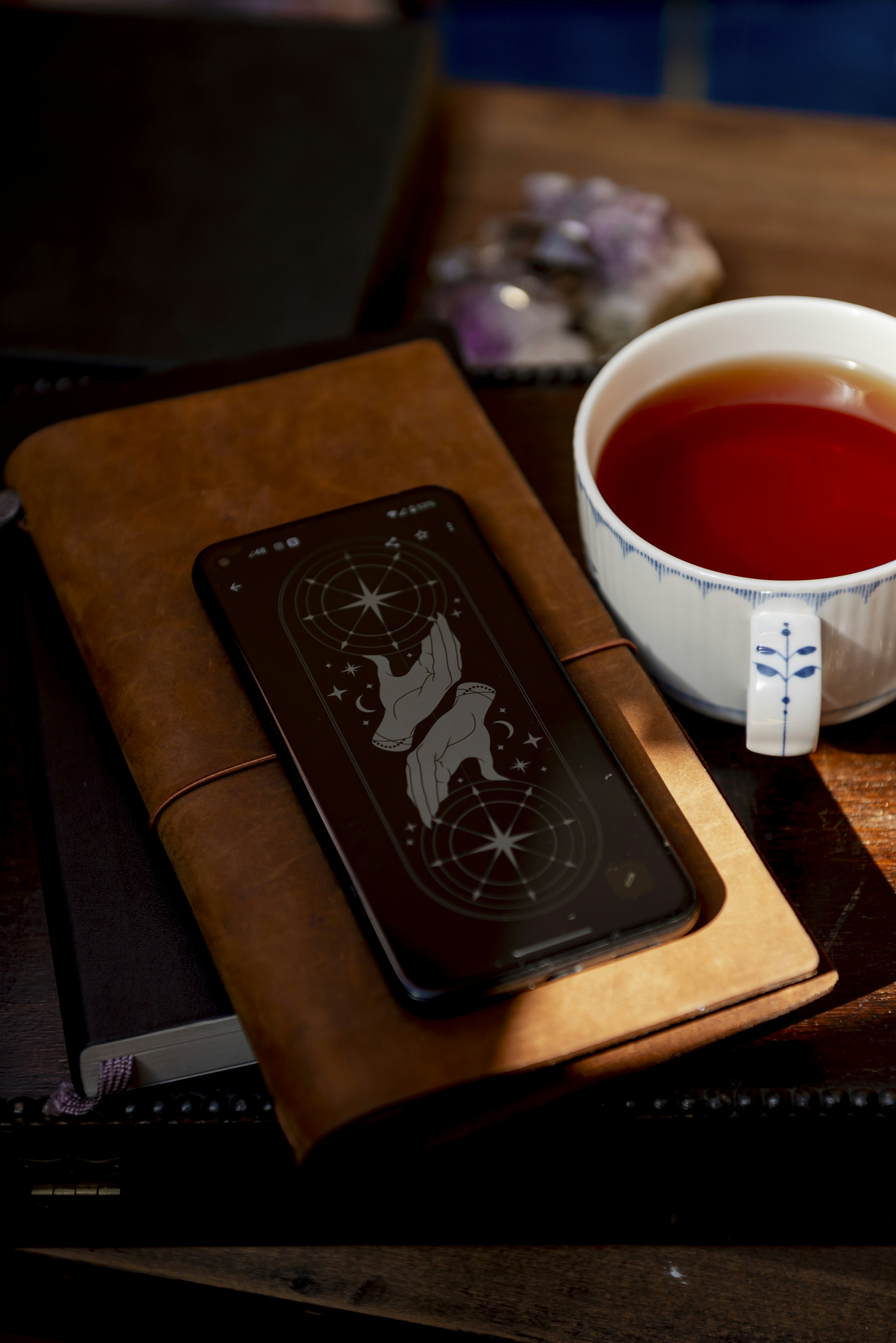 Smartphone displaying tarot-inspired digital wallpaper rests on a leather notebook beside a cup of tea.
