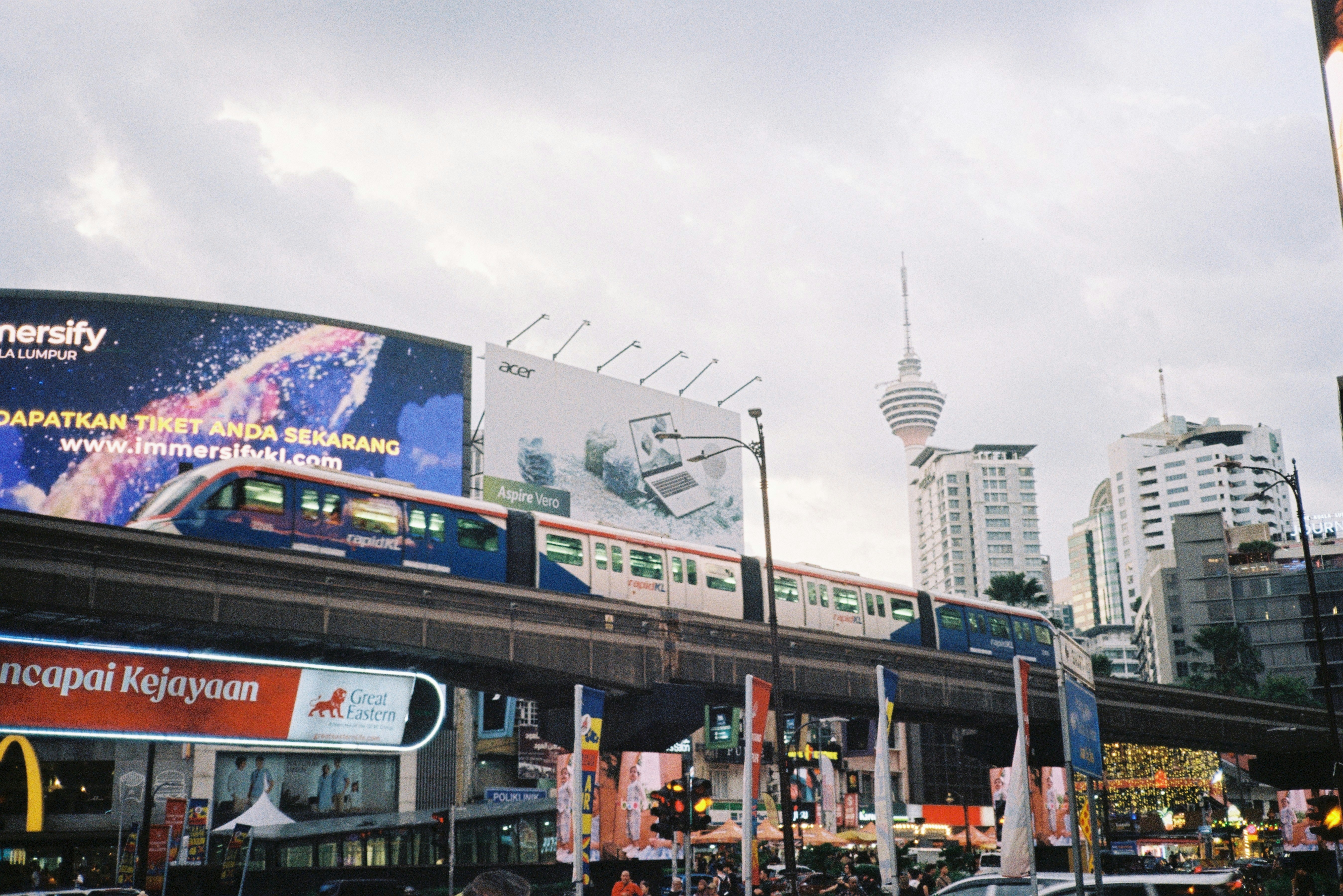 Monorail gliding above bustling Kuala Lumpur streets with skyscrapers and billboards in the background.