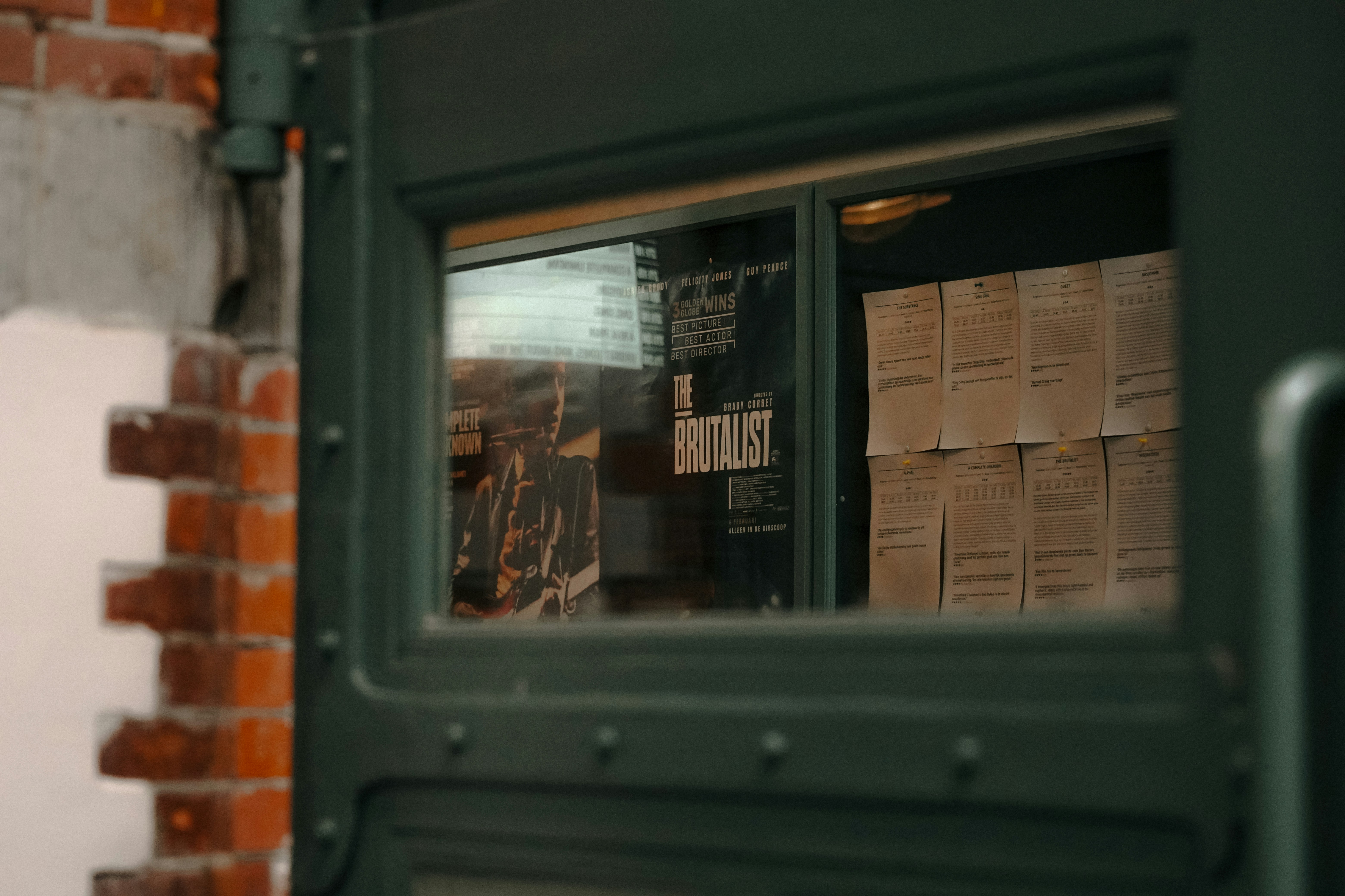A window displaying a film poster for 'The Brutalist' alongside scattered notes, framed by a rustic green door. The scene hints at a narrative waiting to unfold.