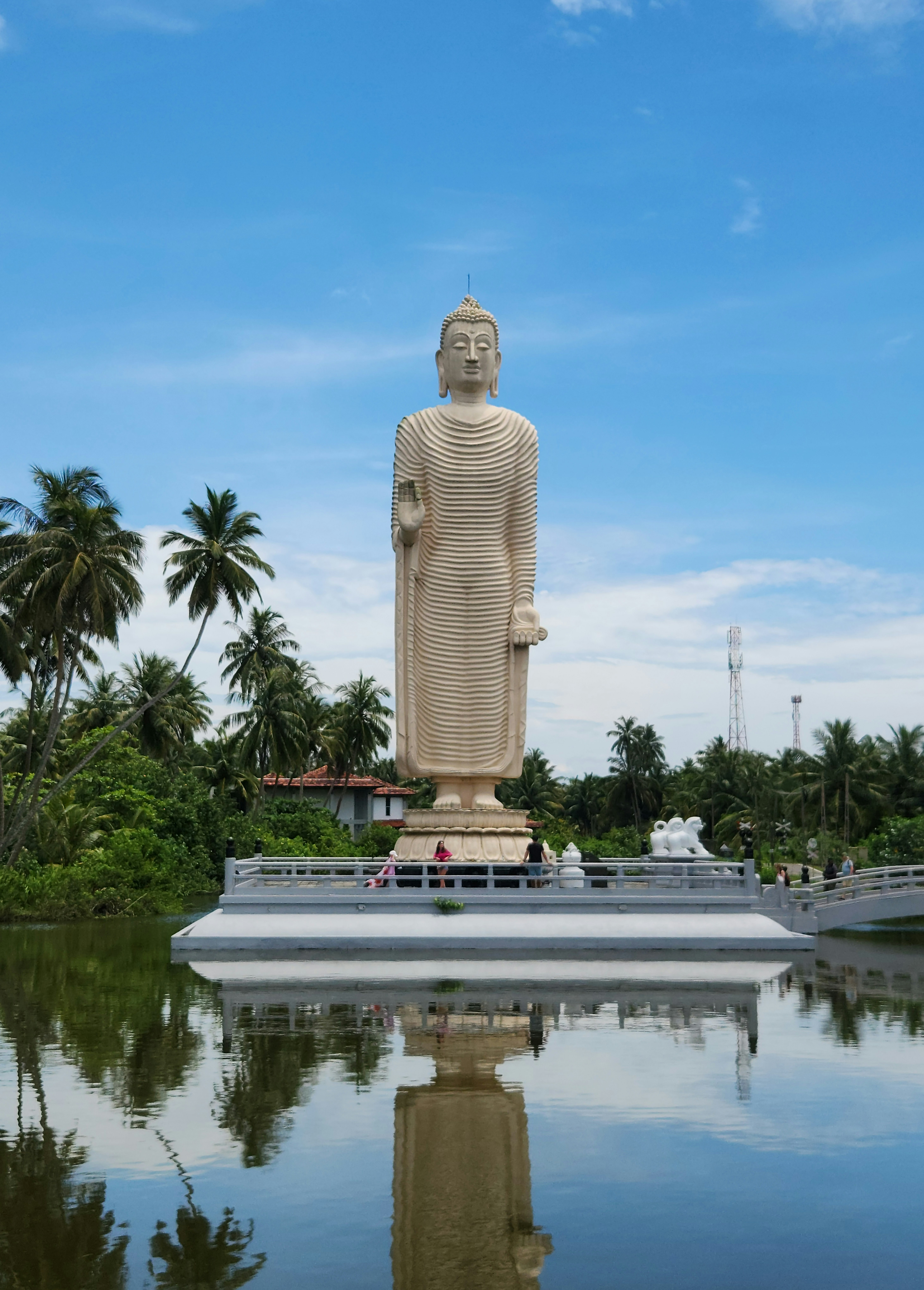Towering Buddha statue on a platform surrounded by water with palm trees and a clear blue sky.