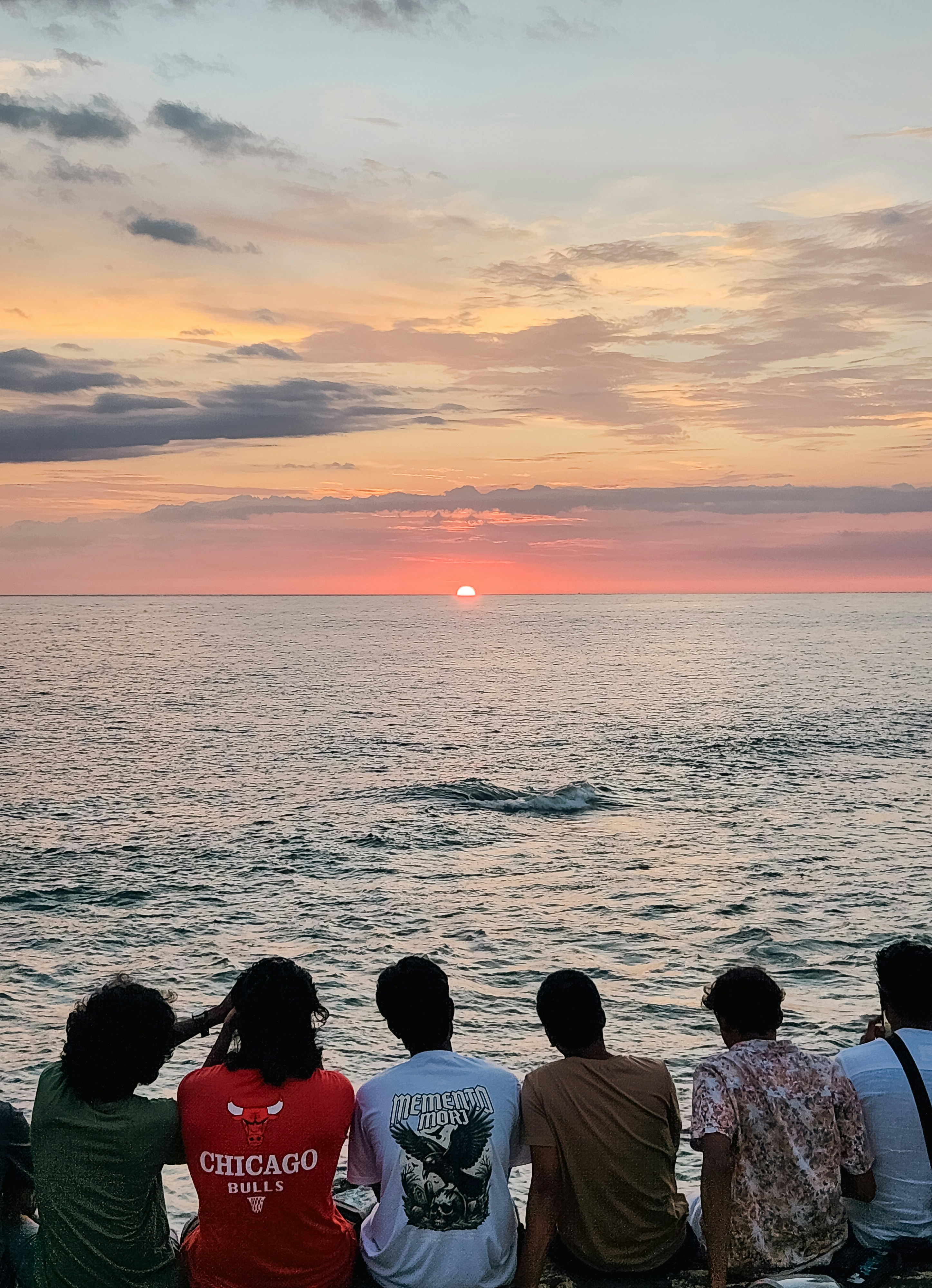 Friends sitting shoulder-to-shoulder on the edge of a cliff looking at the sun set into the Indian Ocean. Note: This photo was clicked at Galle Fort, Sri Lanka with the consent of the entire friend group.