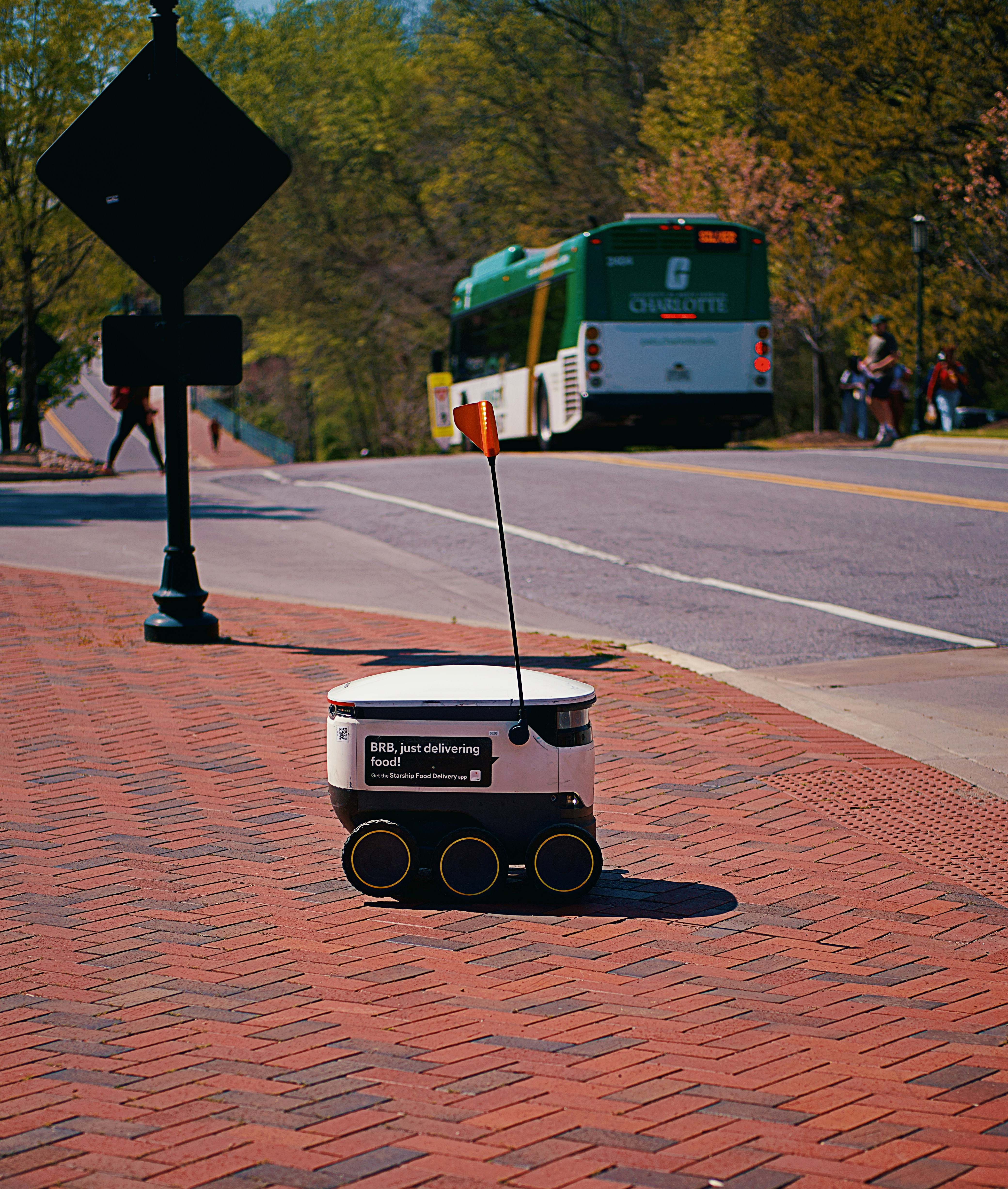 Autonomous delivery robot on a brick path near a road with a bus in the background.