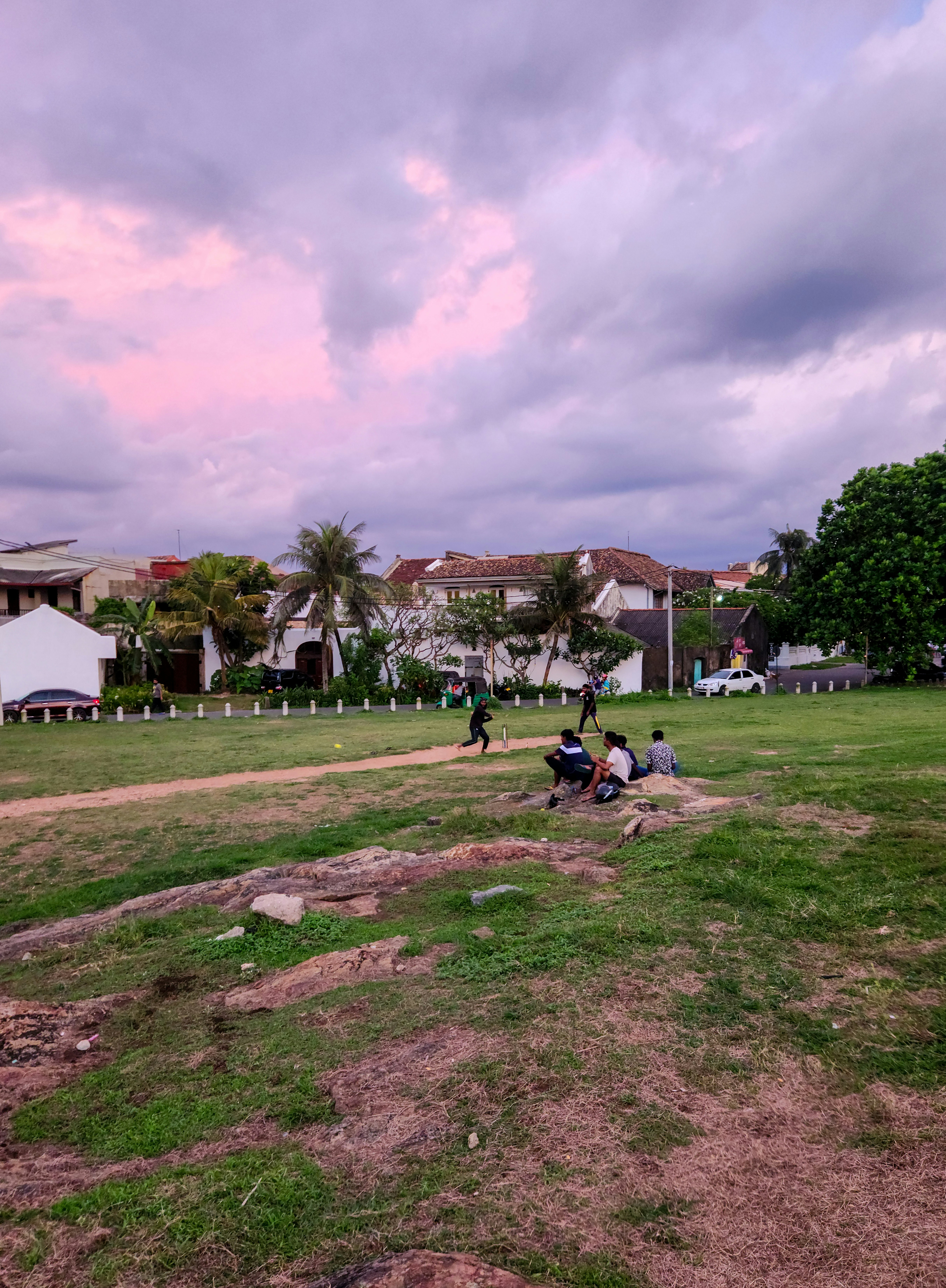 People relaxing on a grassy field under a purple-hued sky at dusk, with colonial buildings in the background.