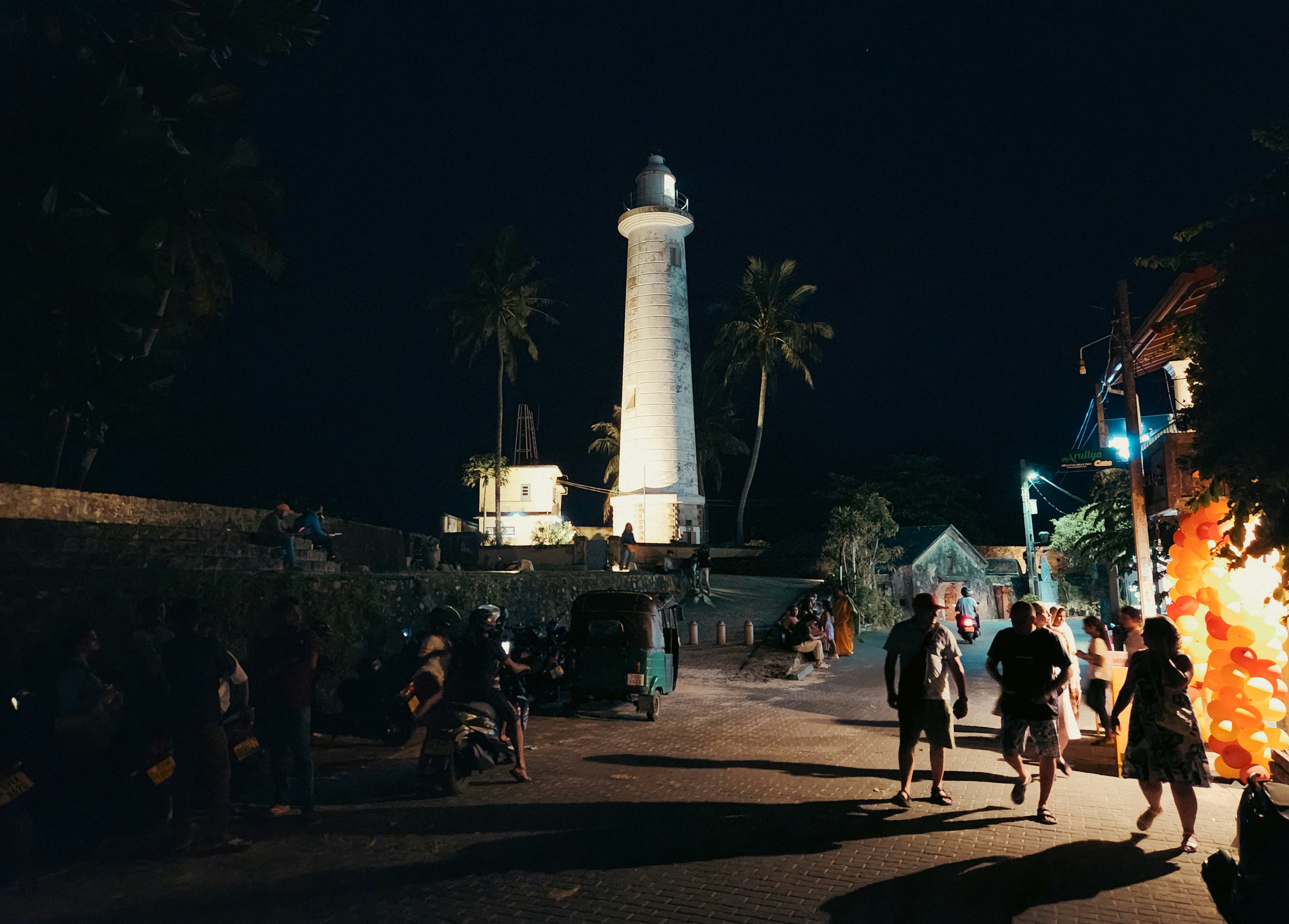 Galle Lighthouse illuminated against the night sky, framed by palm trees and bustling street activity.