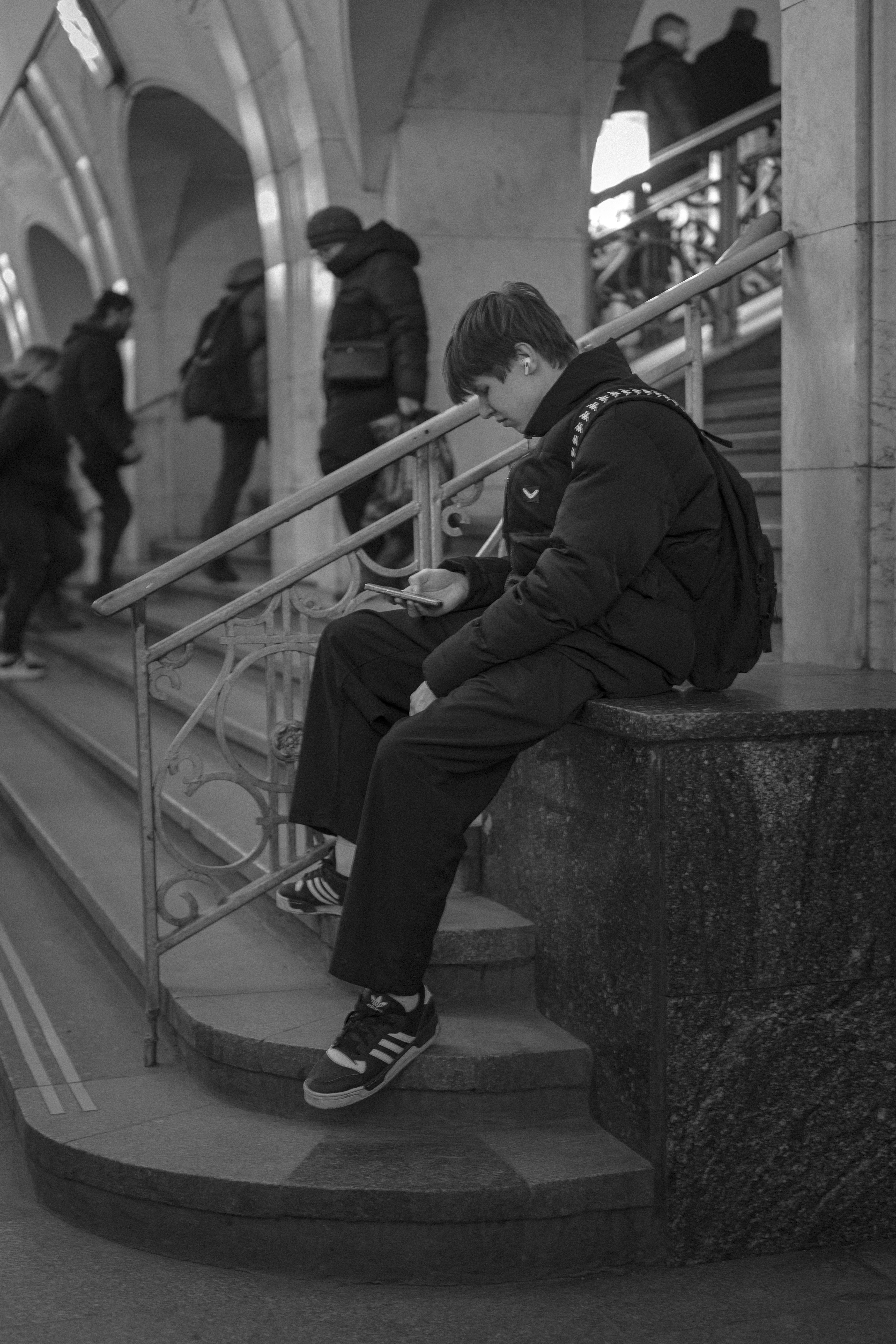 Young man engrossed in his smartphone while seated on steps in a bustling Moscow Metro station.