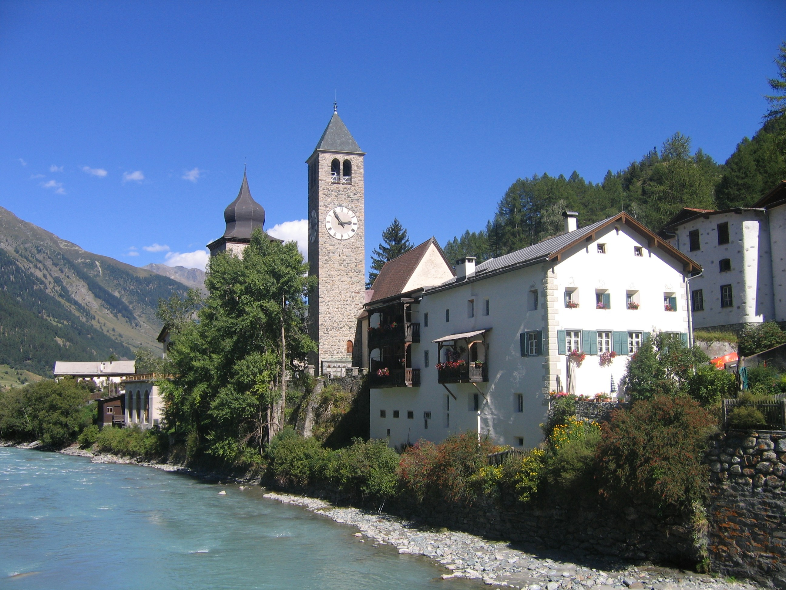 Historic village buildings and clock tower alongside a tranquil river under a clear blue sky.