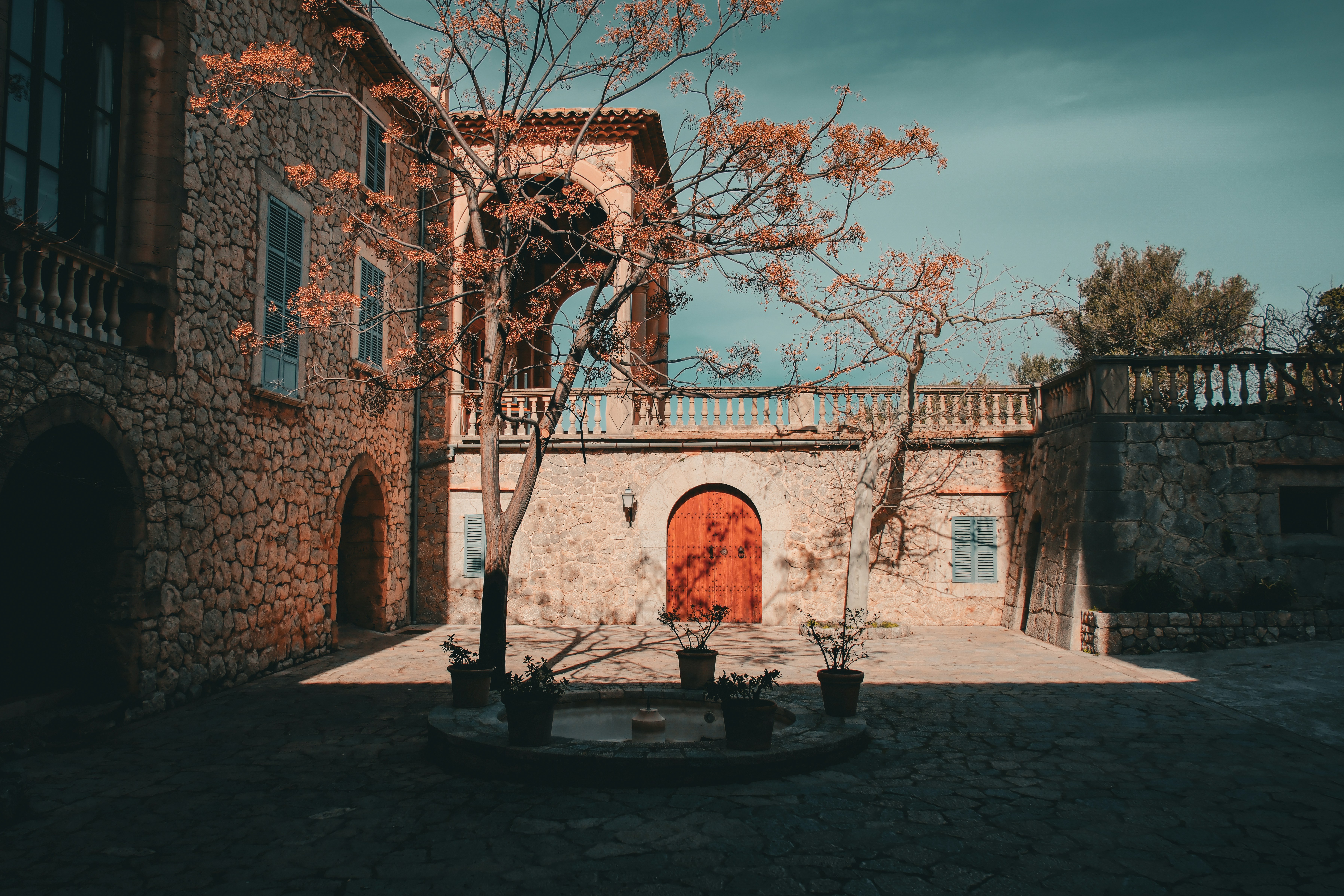 Sun-dappled courtyard with a vibrant red door, surrounded by rustic stone architecture and a leafless tree.