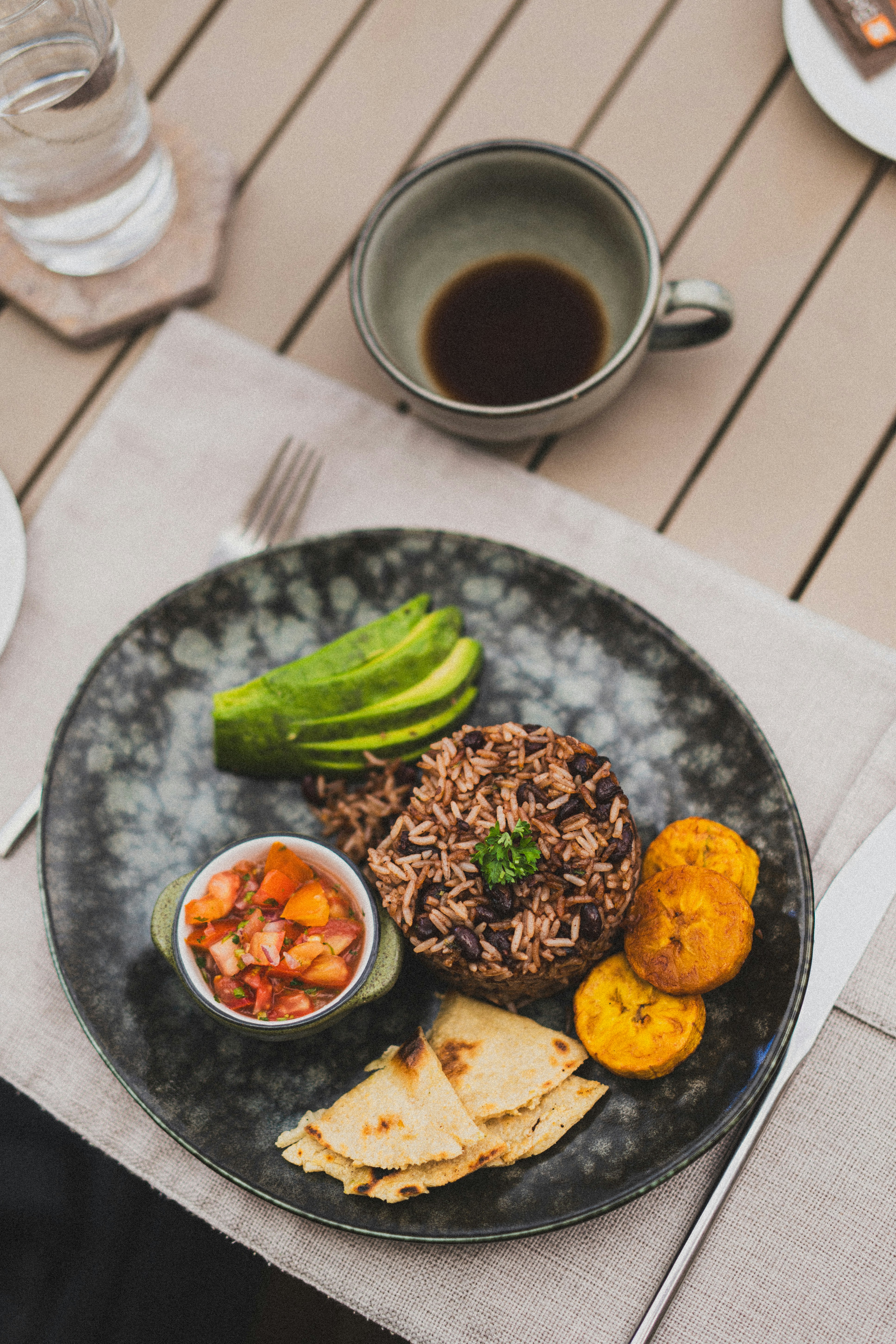 A vibrant plate featuring rice and beans, ripe avocado slices, fried plantains, and a fresh salsa. The dish is artfully arranged on a textured plate, inviting a culinary experience.