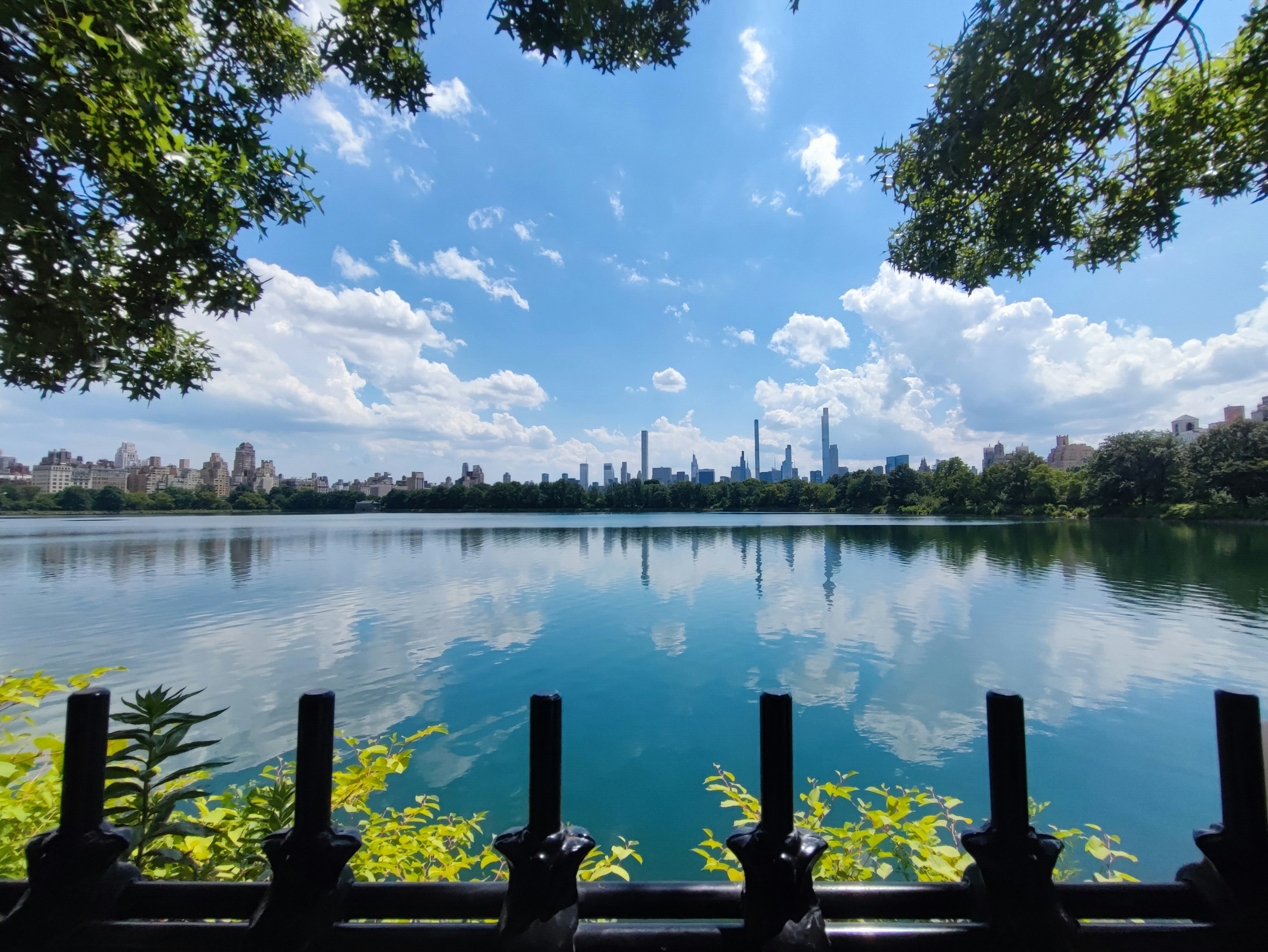 City skyline mirrored on a serene lake, framed by leafy trees and a vibrant blue sky with clouds.