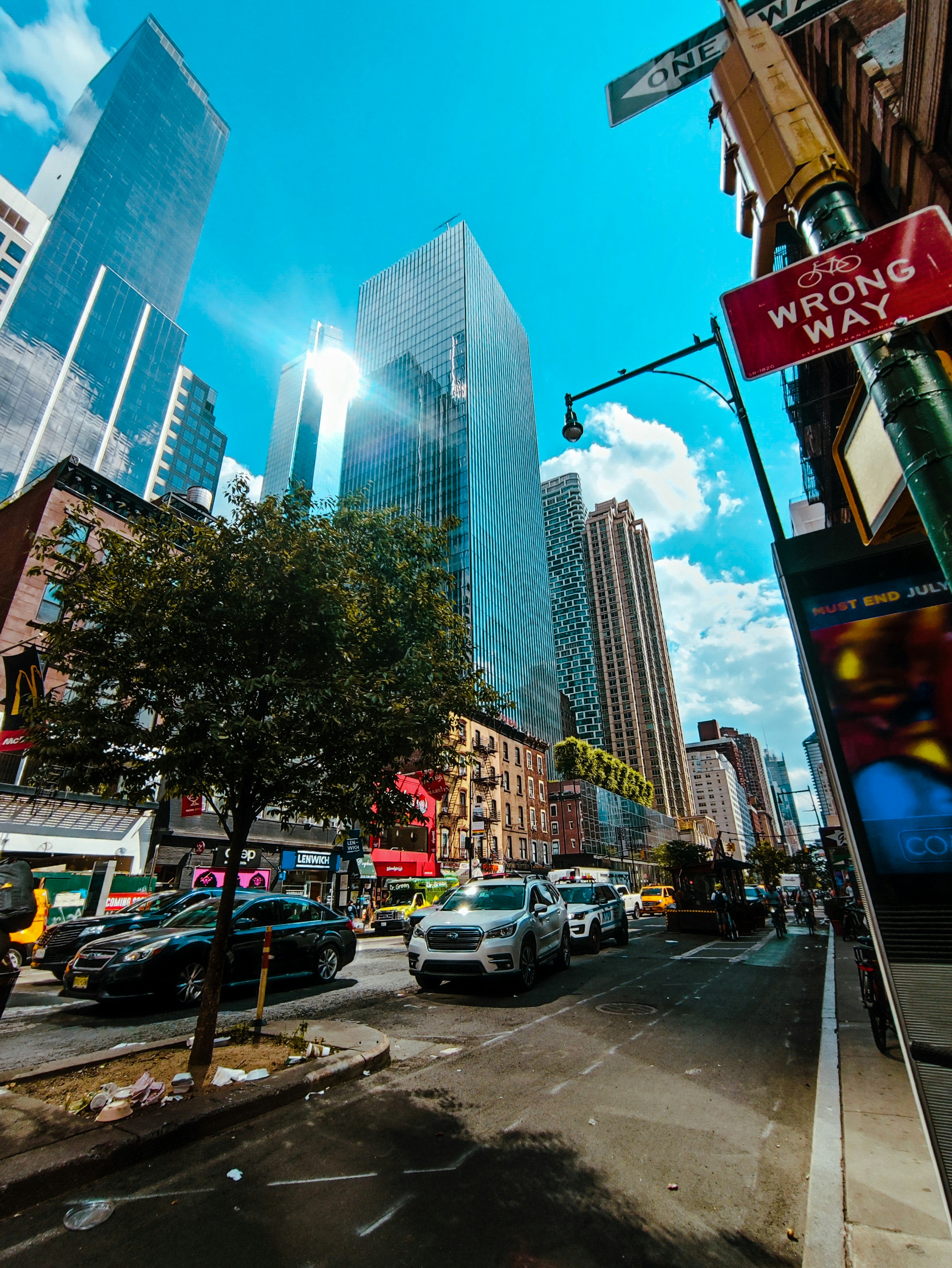 Vibrant city street bustling with traffic and pedestrians, framed by towering skyscrapers under a bright blue sky.