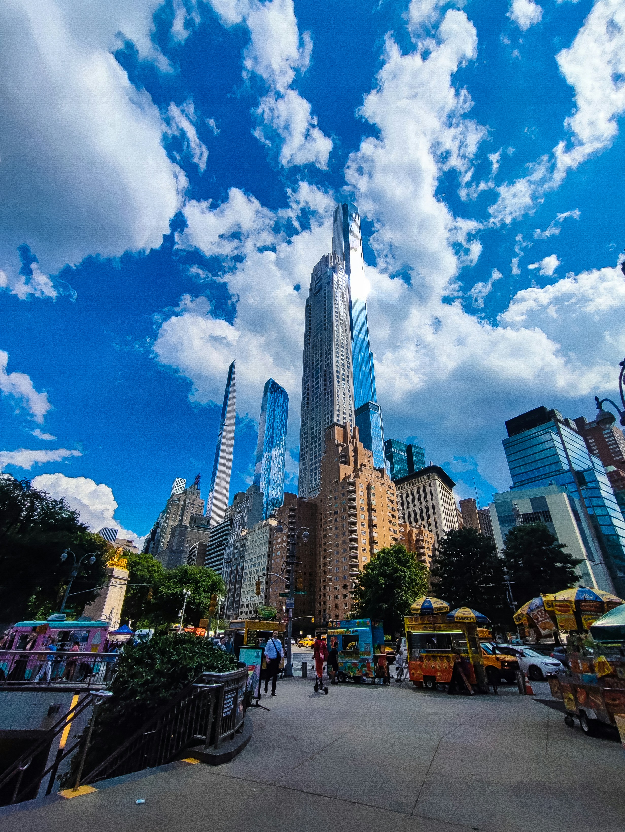 Towering skyscrapers rise against a vibrant blue sky with scattered clouds, bustling street below with pedestrians and vendors.