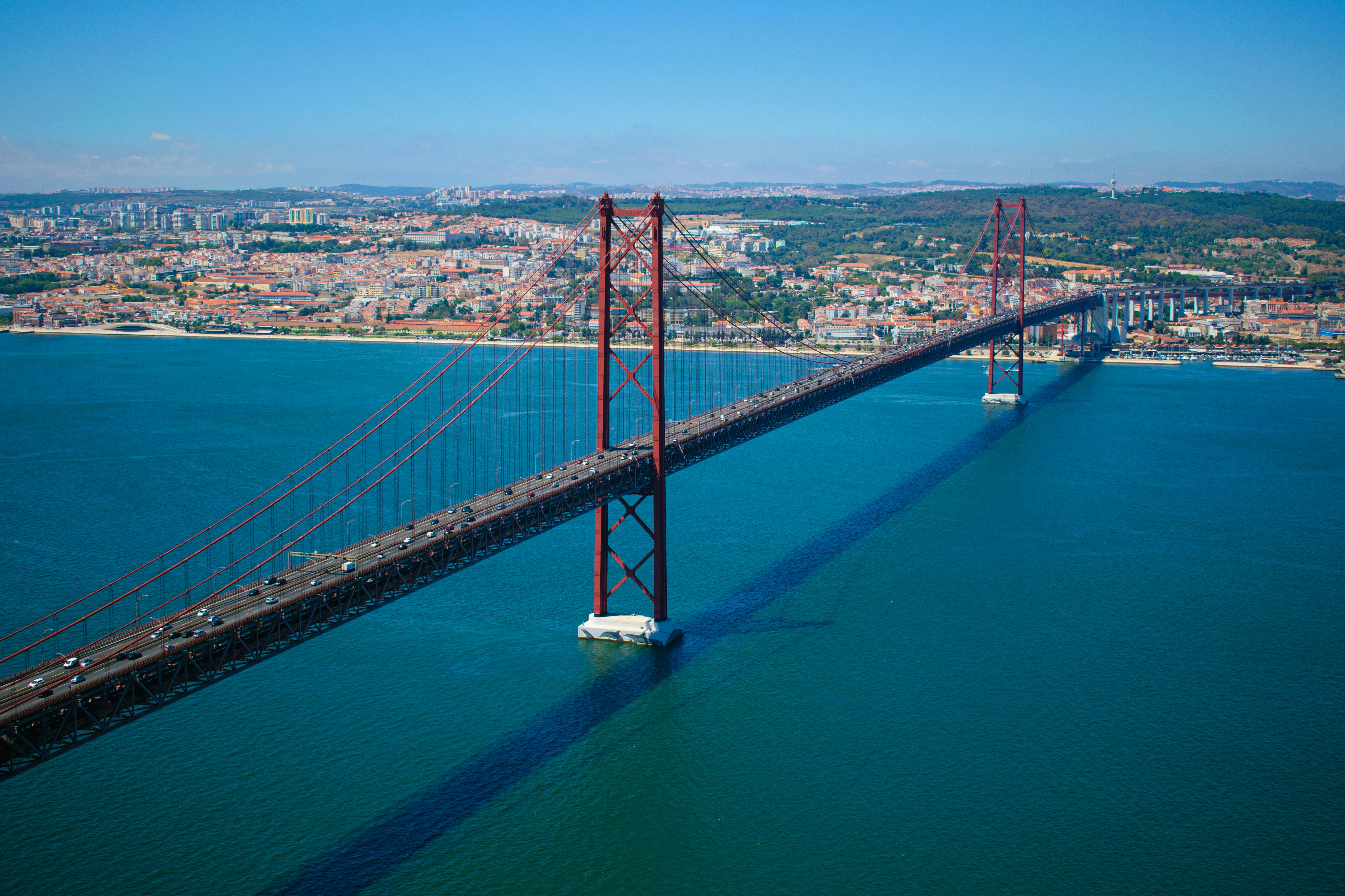 Aerial view of the 25th of April Bridge stretching across Lisbon's water under a clear blue sky.