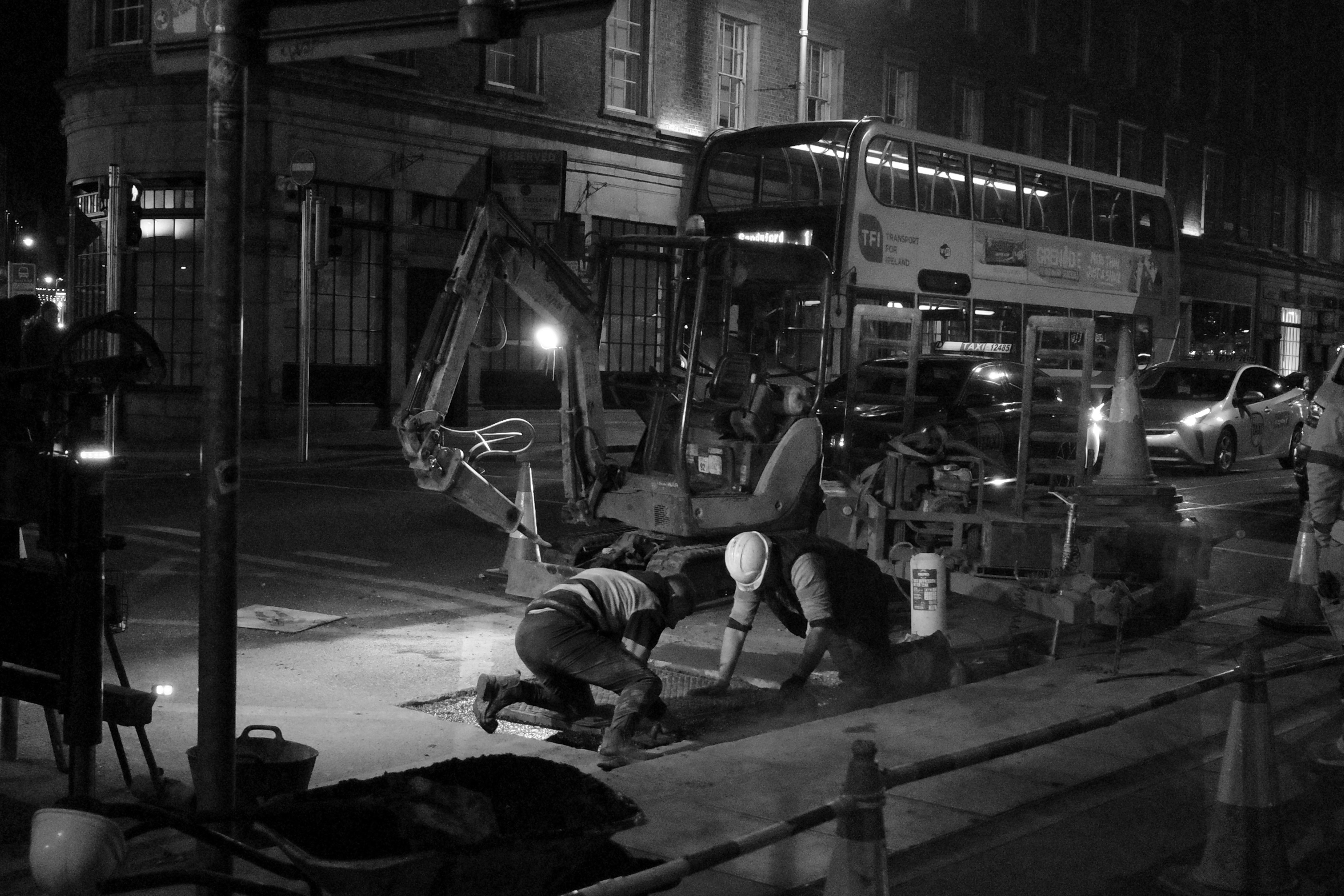 Workers engaged in nighttime street construction with machinery and a bus in the background.
