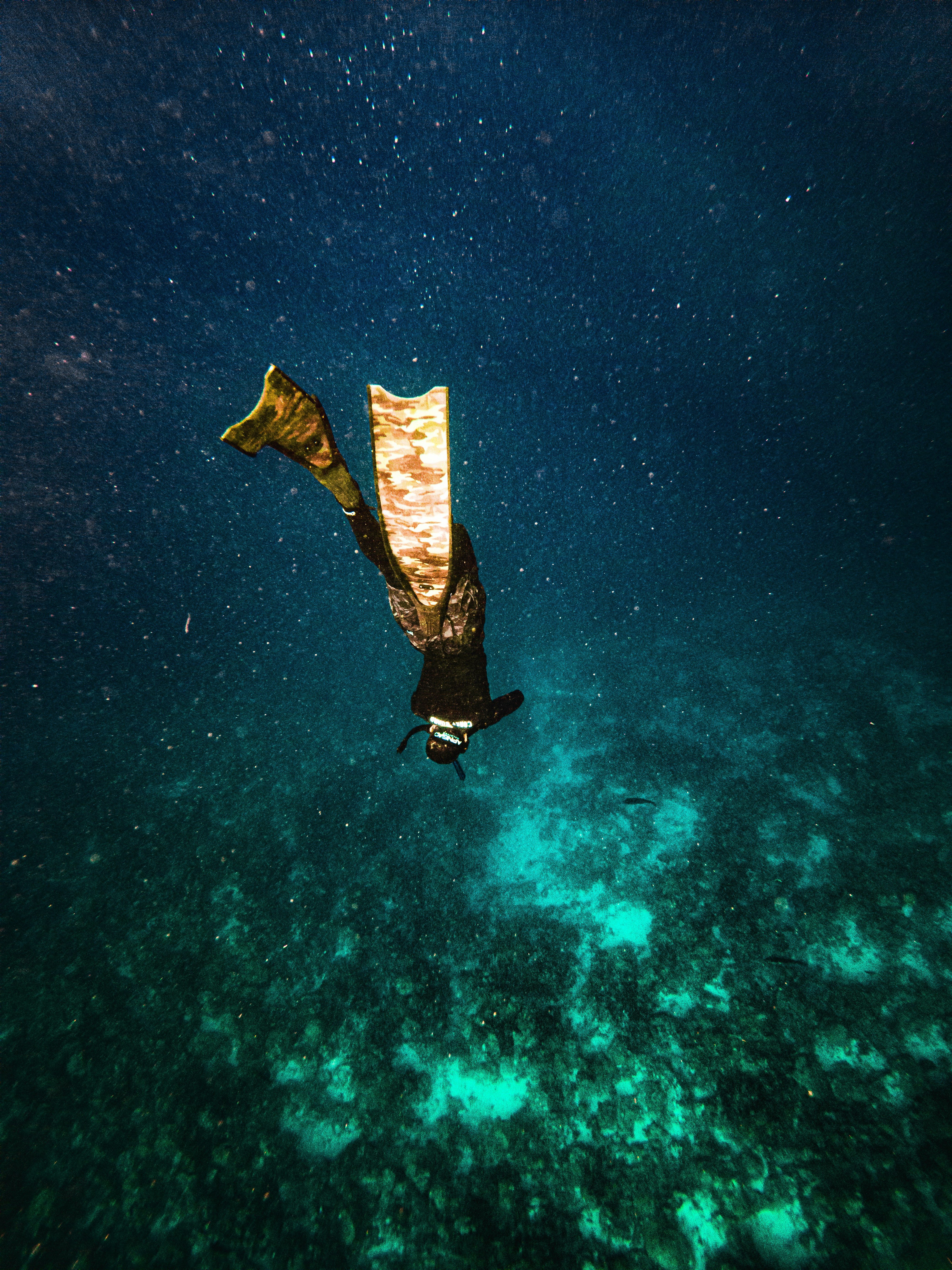 A lone freediver descends into deep blue, speckled water. Fins pierce the darkness, hinting at the rocky seabed below. A moment of serene isolation in the vast ocean's embrace.