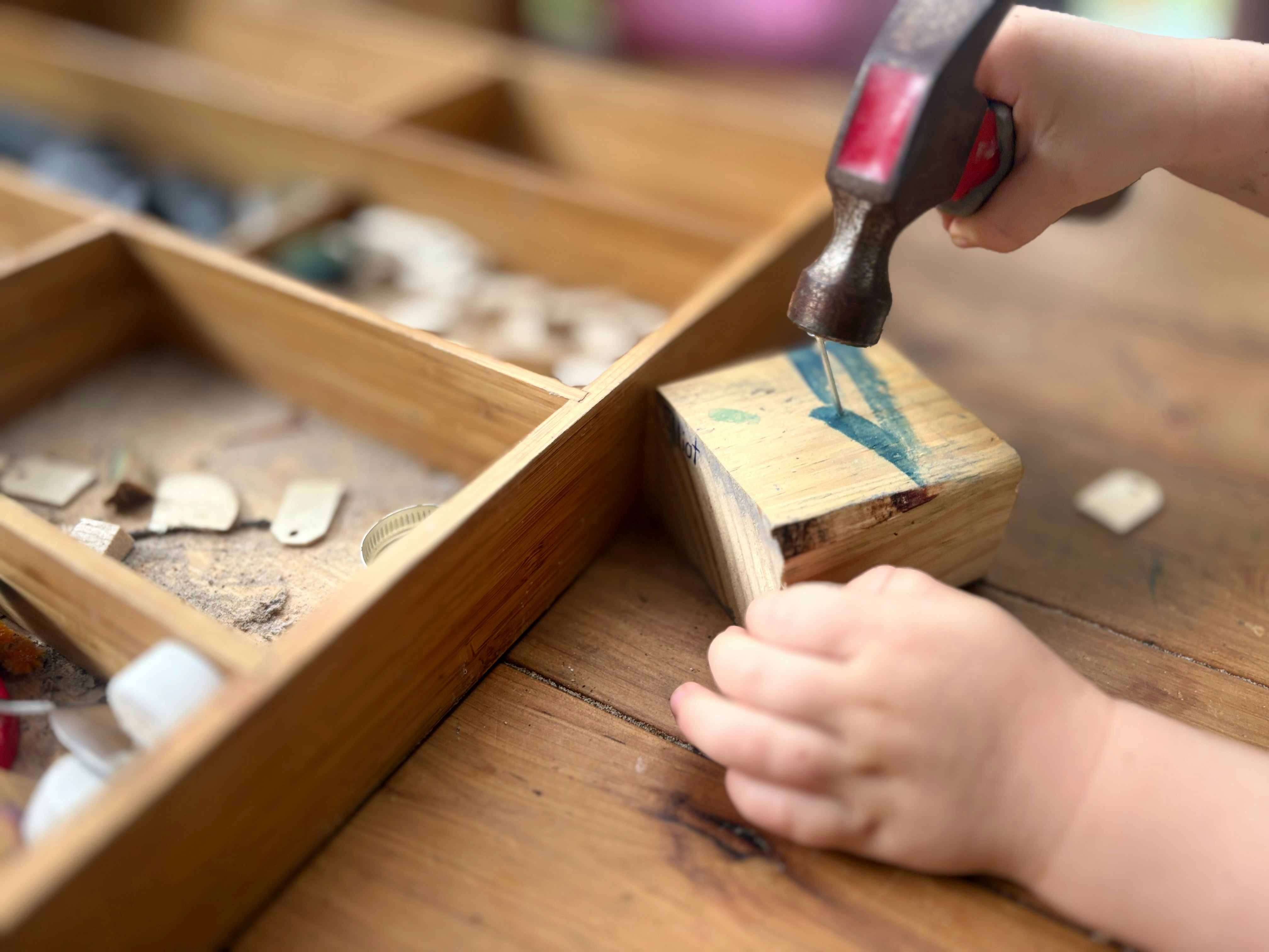 Child's hand holding a hammer, striking a wooden block adorned with a painted design, surrounded by various crafting materials in a wooden tray.