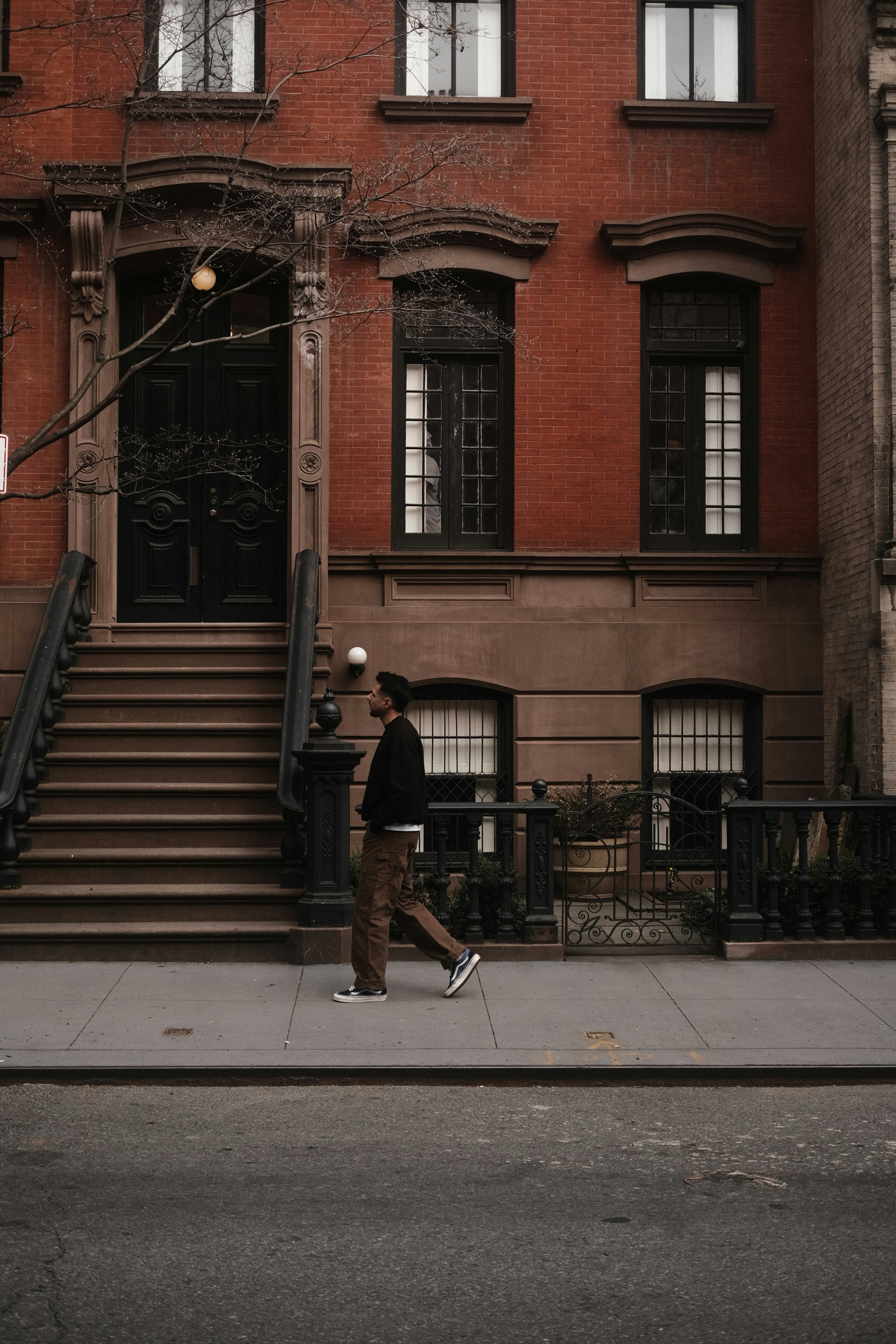 Person walking on a sidewalk past a classic red brick townhouse with ornate details and steps in low light.