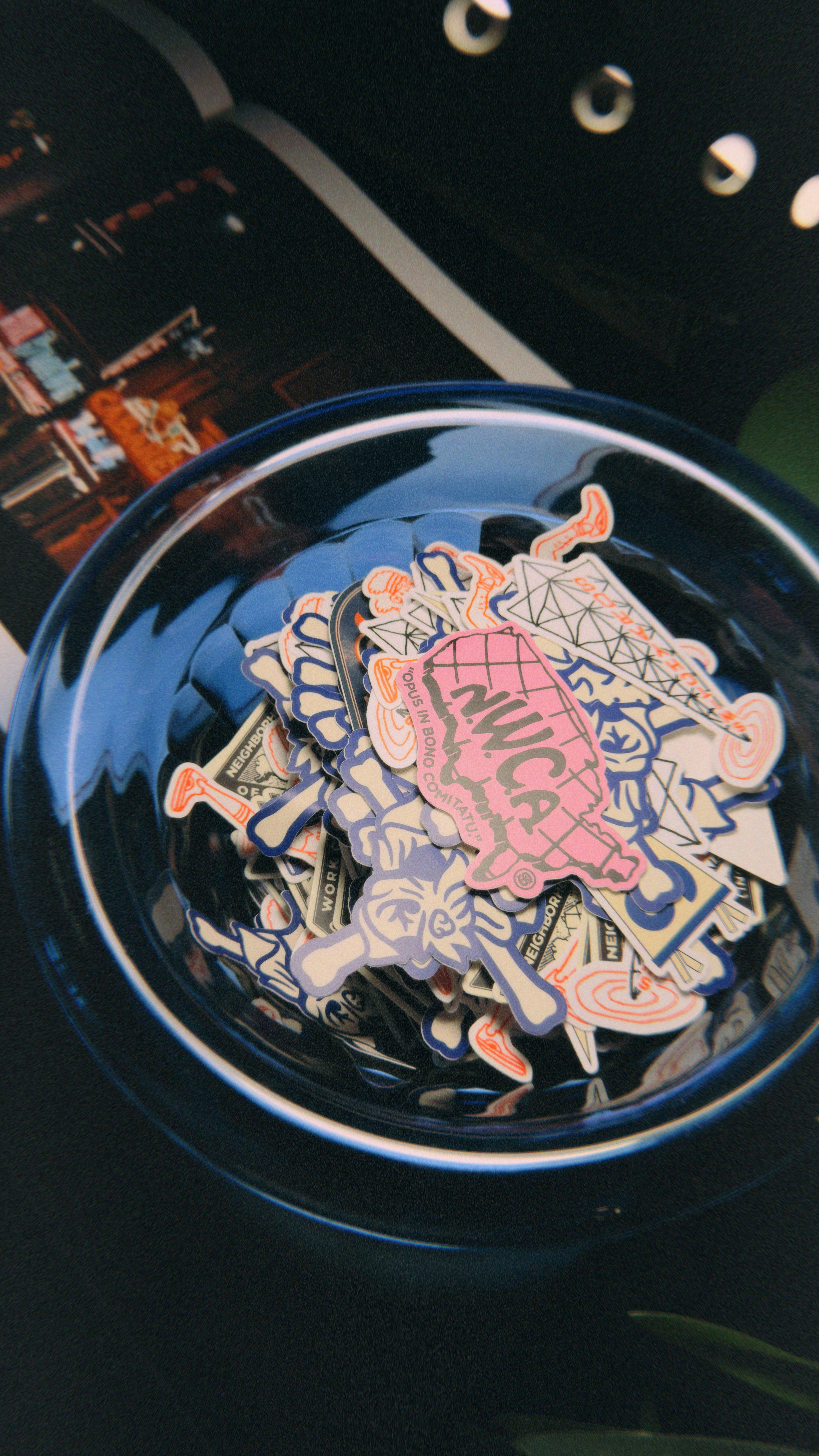 Colorful assortment of stickers in a blue dish, showcasing playful designs and vibrant hues. The background features a glimpse of a magazine page.