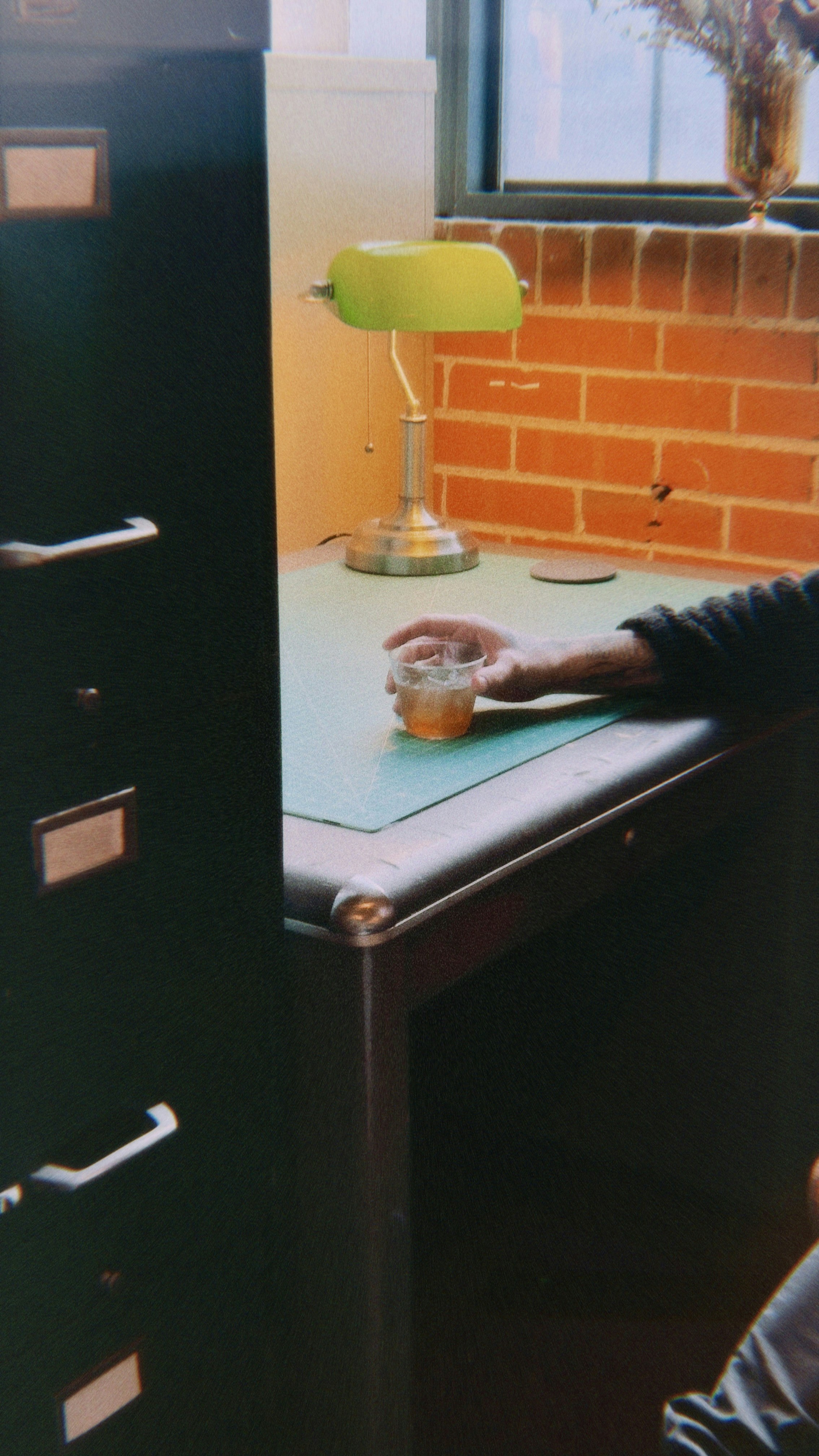 A hand rests on a desk holding a cup of drink, with a green desk lamp illuminating the workspace. The backdrop features a brick wall, adding warmth to the scene.