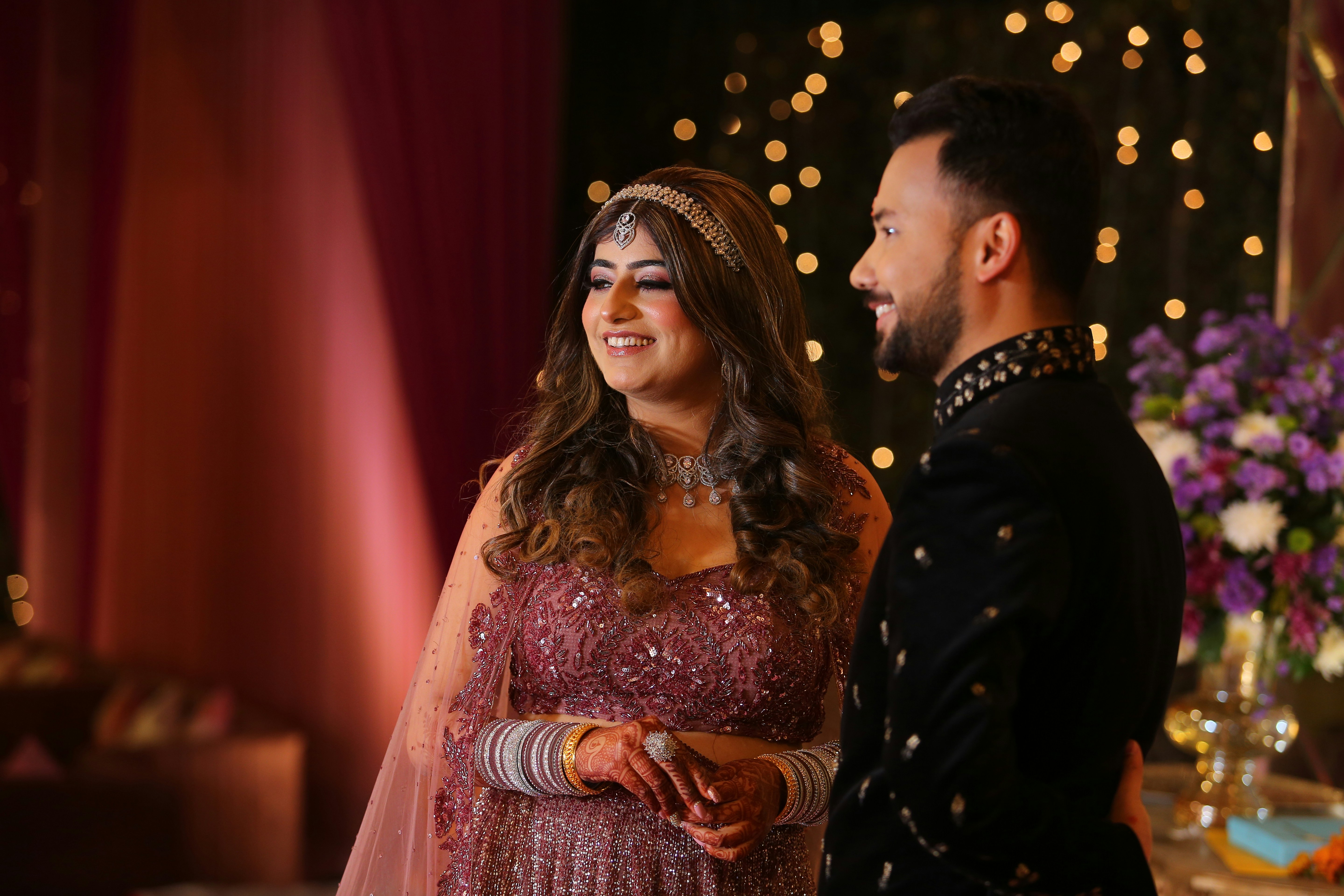 Smiling couple in elegant attire, surrounded by warm lighting and floral decorations.