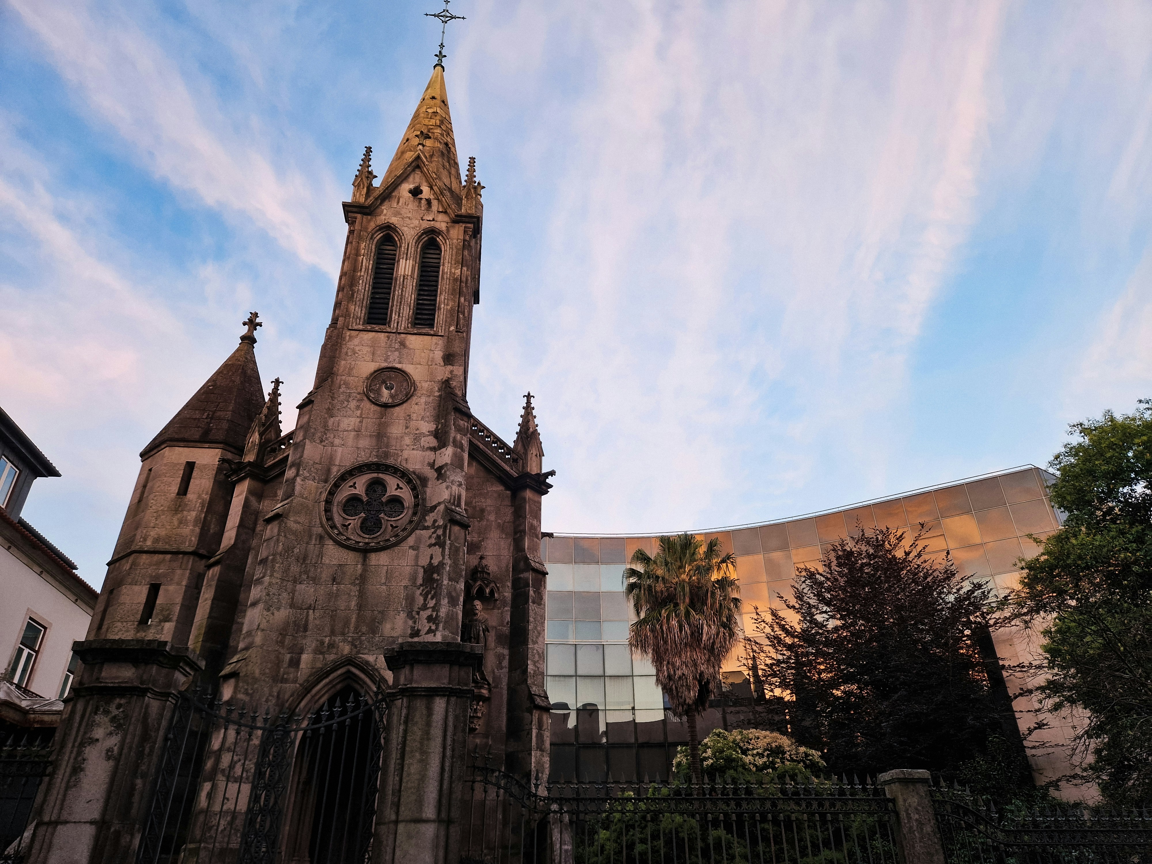 Gothic church tower juxtaposed with modern glass building under a pastel sky.
