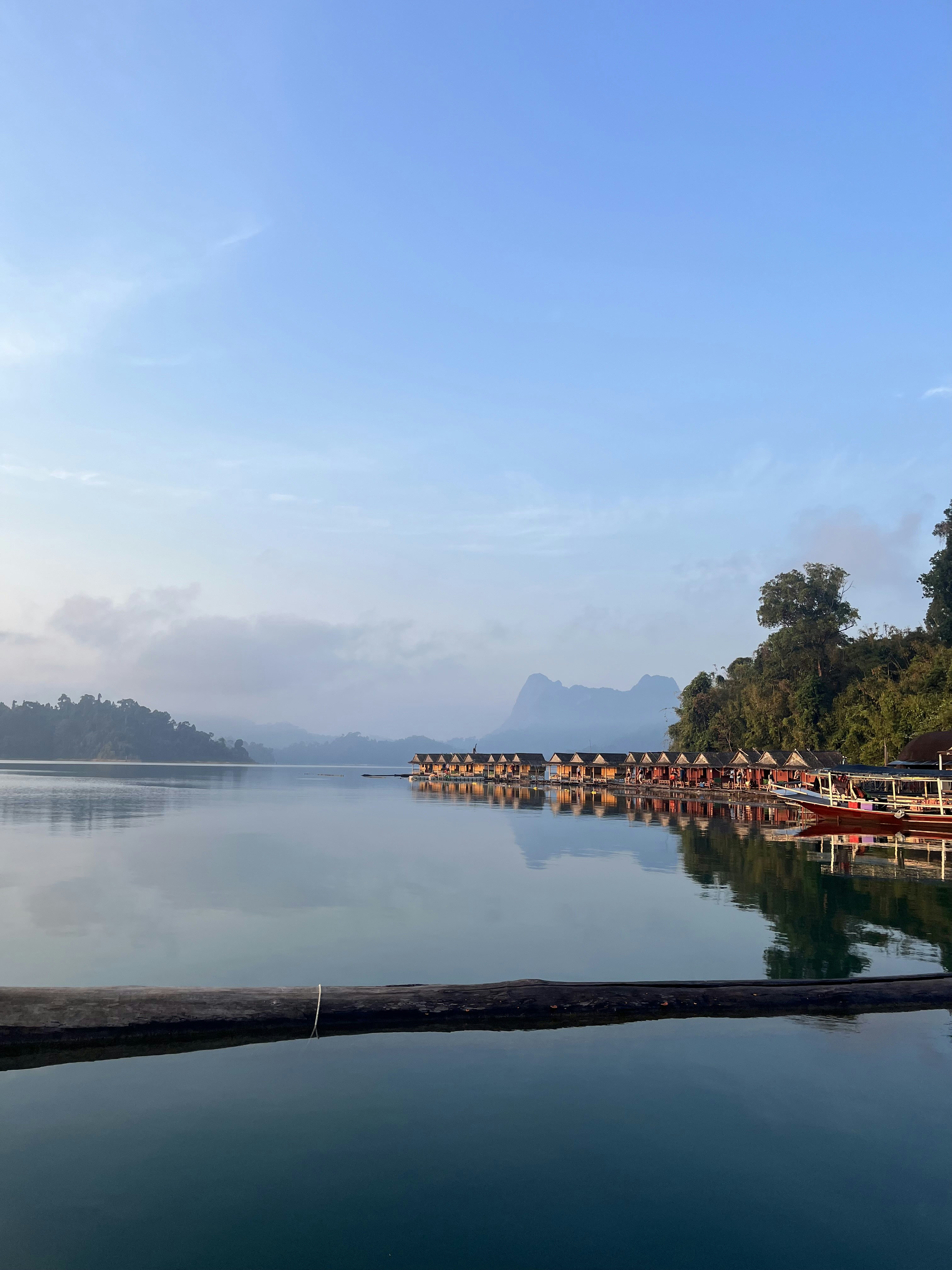 Floating bungalows line a calm lake under a clear blue sky with distant mountains.